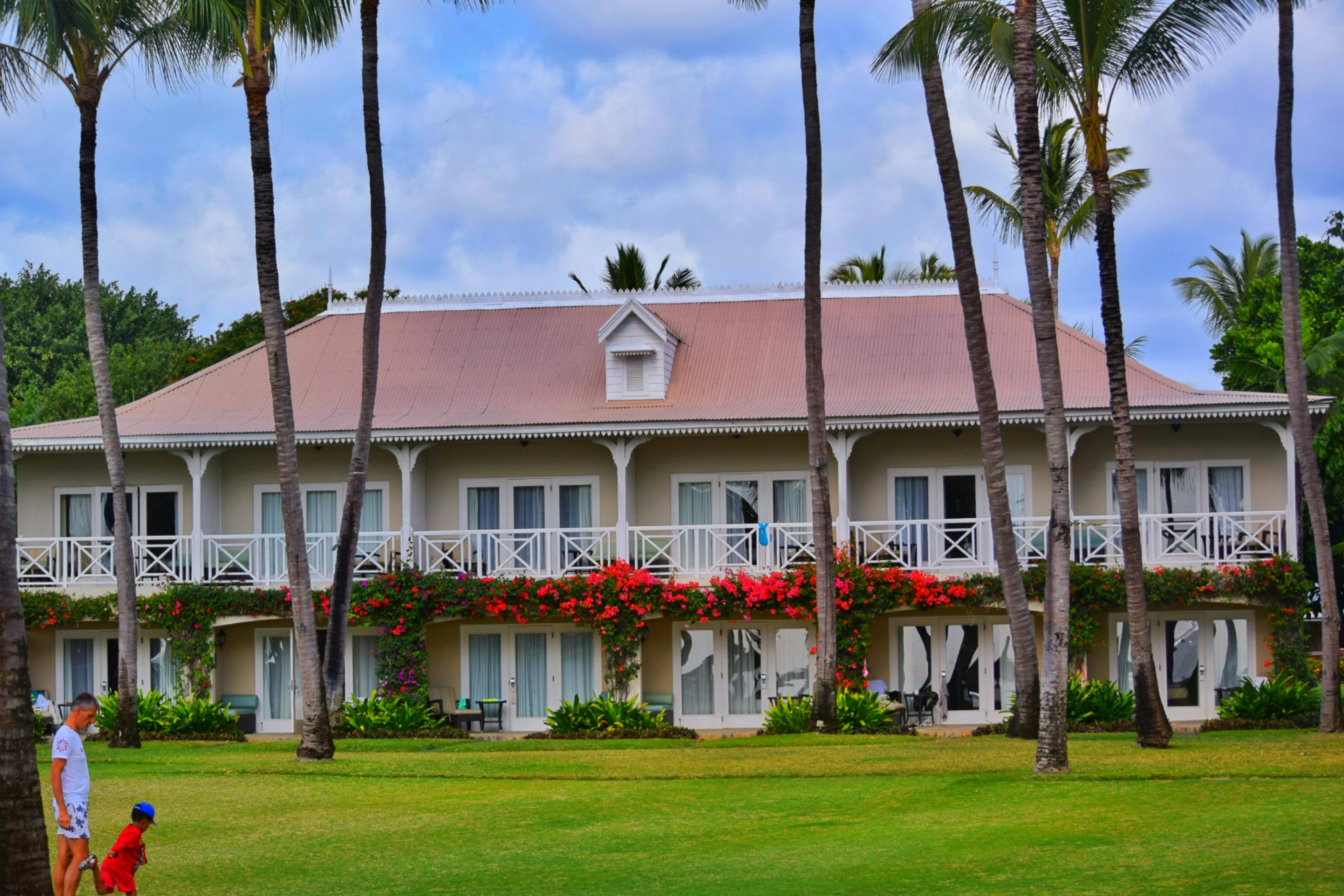 Charming two-story building adorned with vibrant flowers and palm trees, set against a clear sky in a tropical landscape.