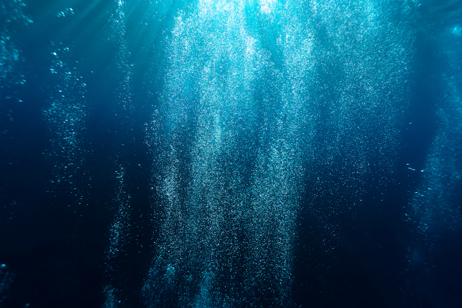 Rays of sunlight filter through deep blue water, illuminating a cloud of rising bubbles. The scene feels like an underwater shaft of light.