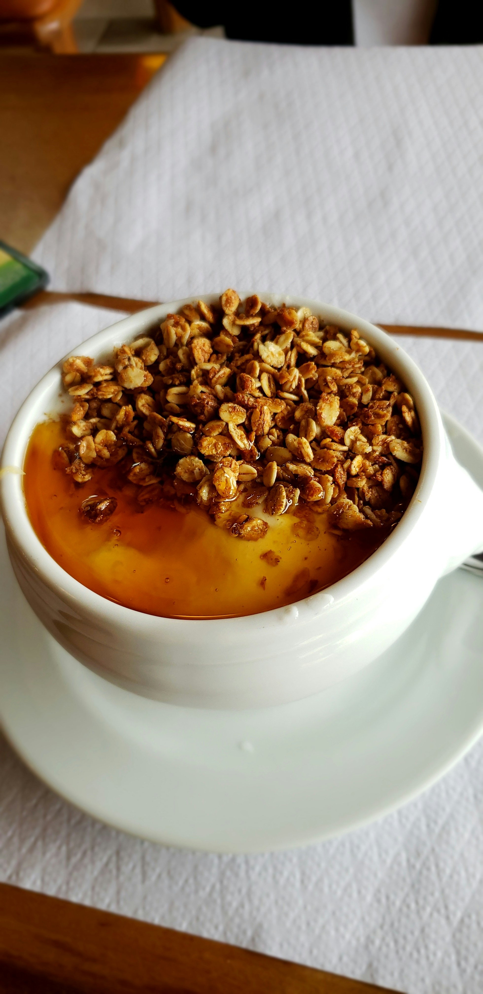 This photograph captures a crème brûlée in a white ramekin topped with a crunchy granola crust, resting on a white saucer with a warm caramel glow.