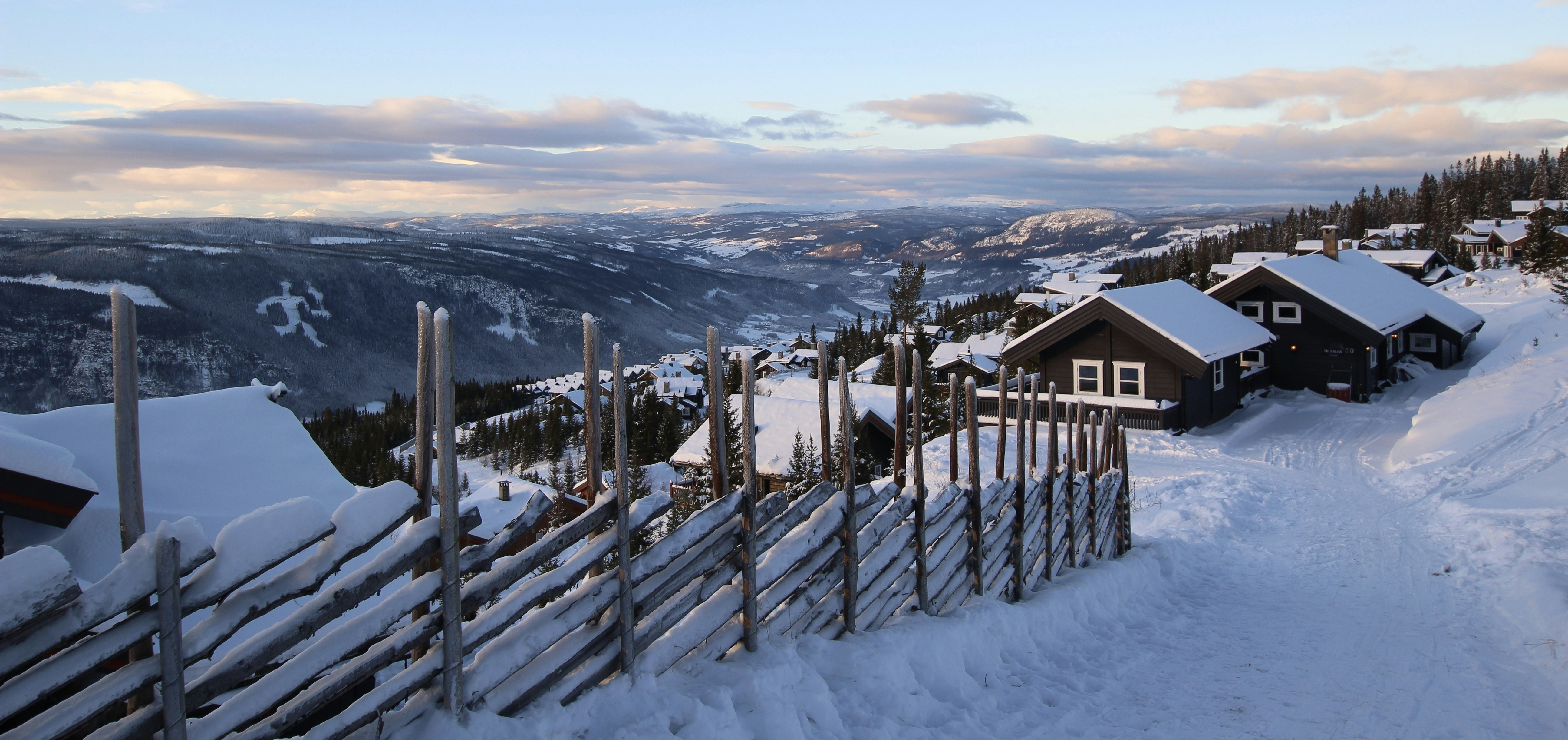 Snow-covered landscape with rustic cabins nestled in the hills, framed by a wooden fence and distant mountains under a soft sky.