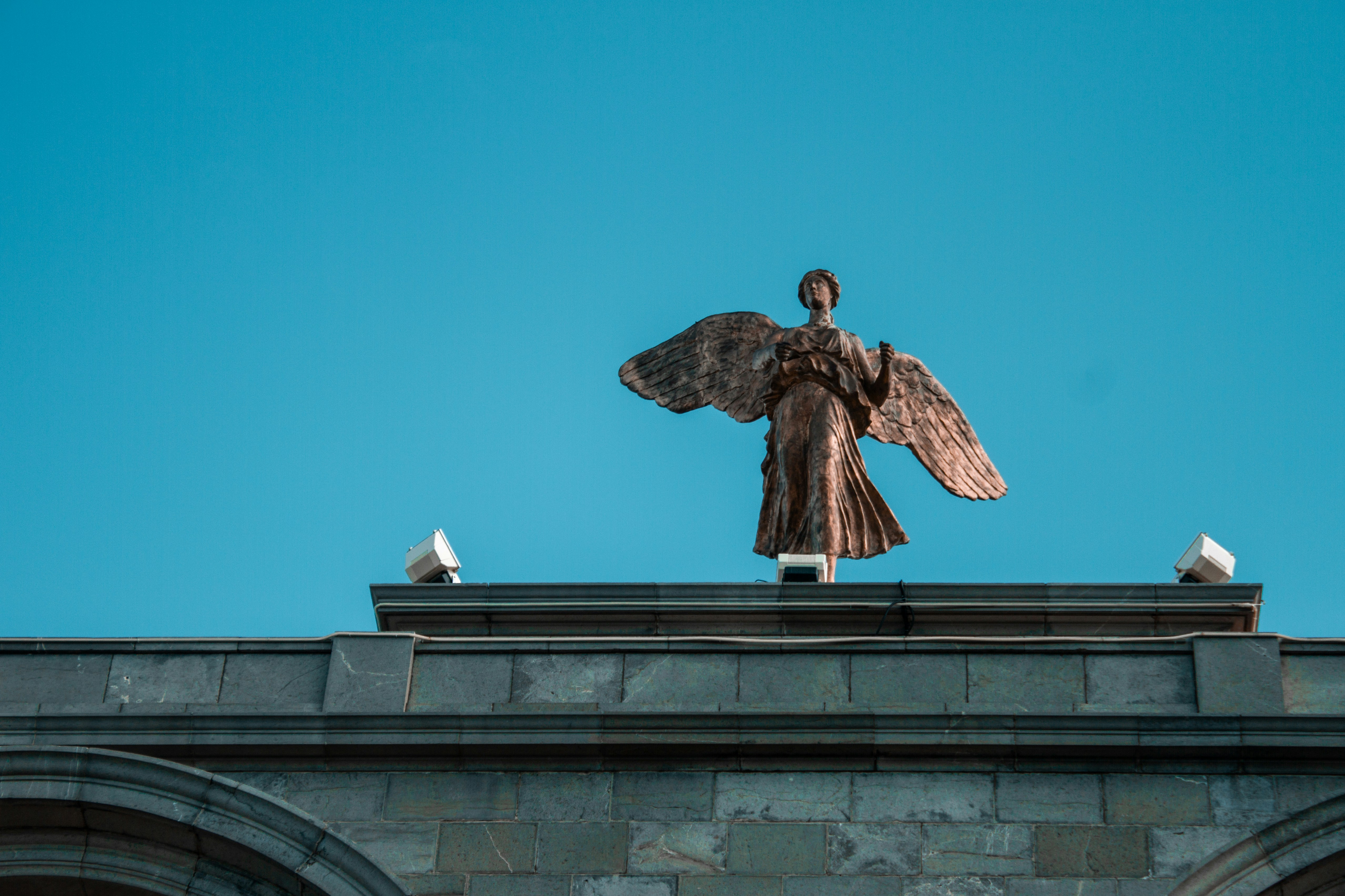 Bronze statue of an angel with outstretched wings atop a stone building under a clear blue sky.