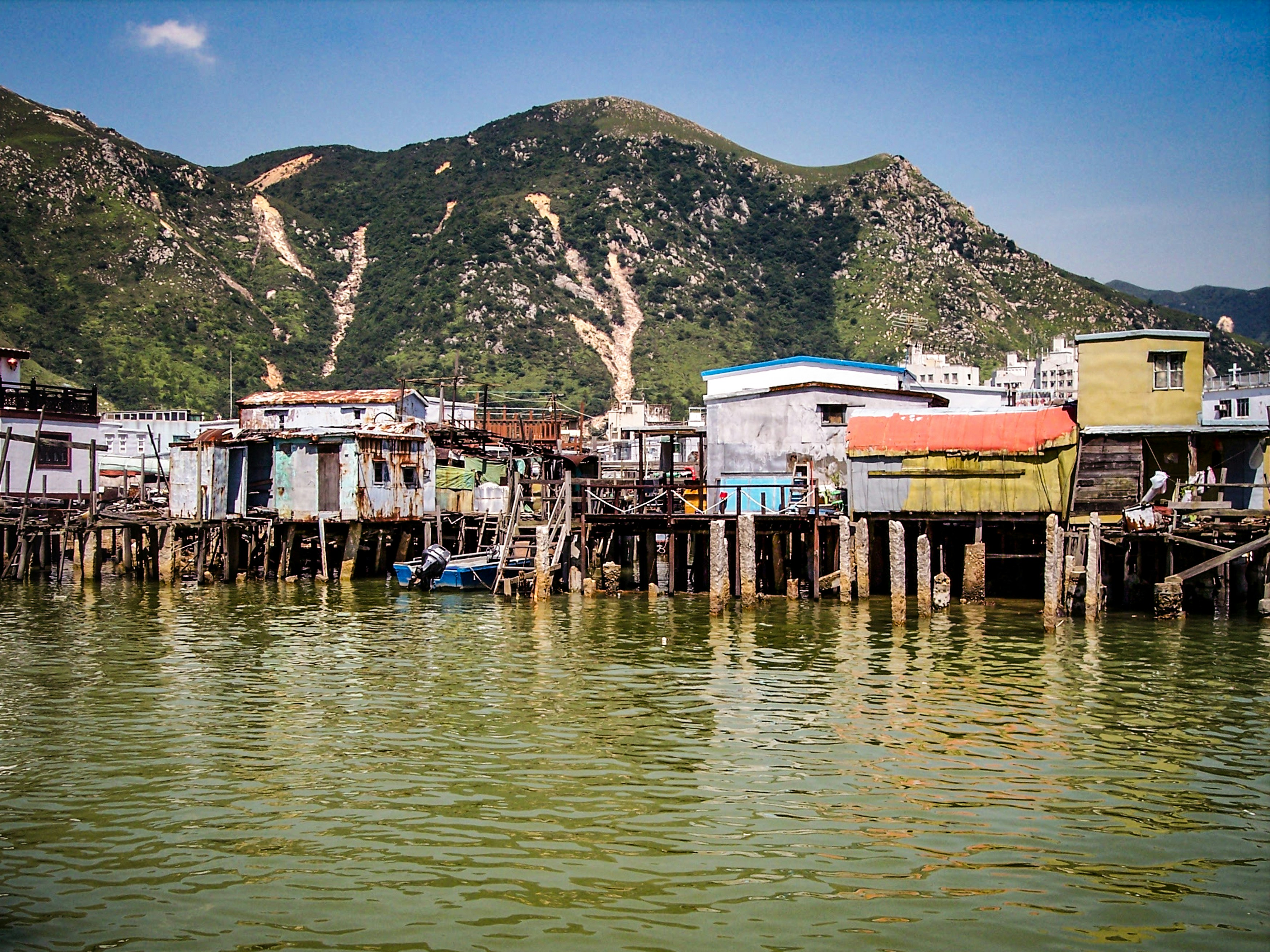 Colorful stilt houses rise above the green waters, framed by lush mountains in the background. The scene captures the essence of a traditional fishing village.