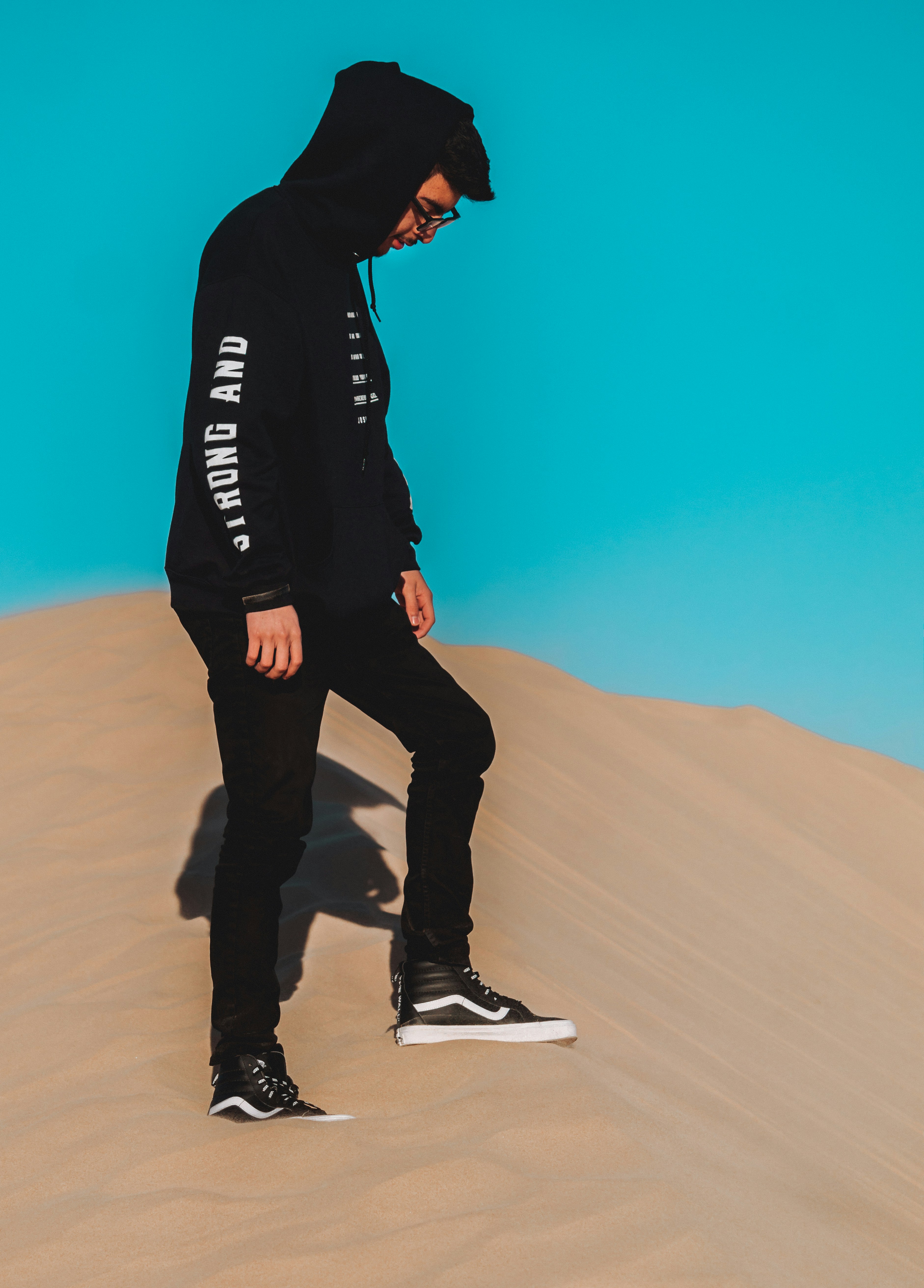 Individual in a black hoodie stands on a sandy dune against a vibrant blue background.