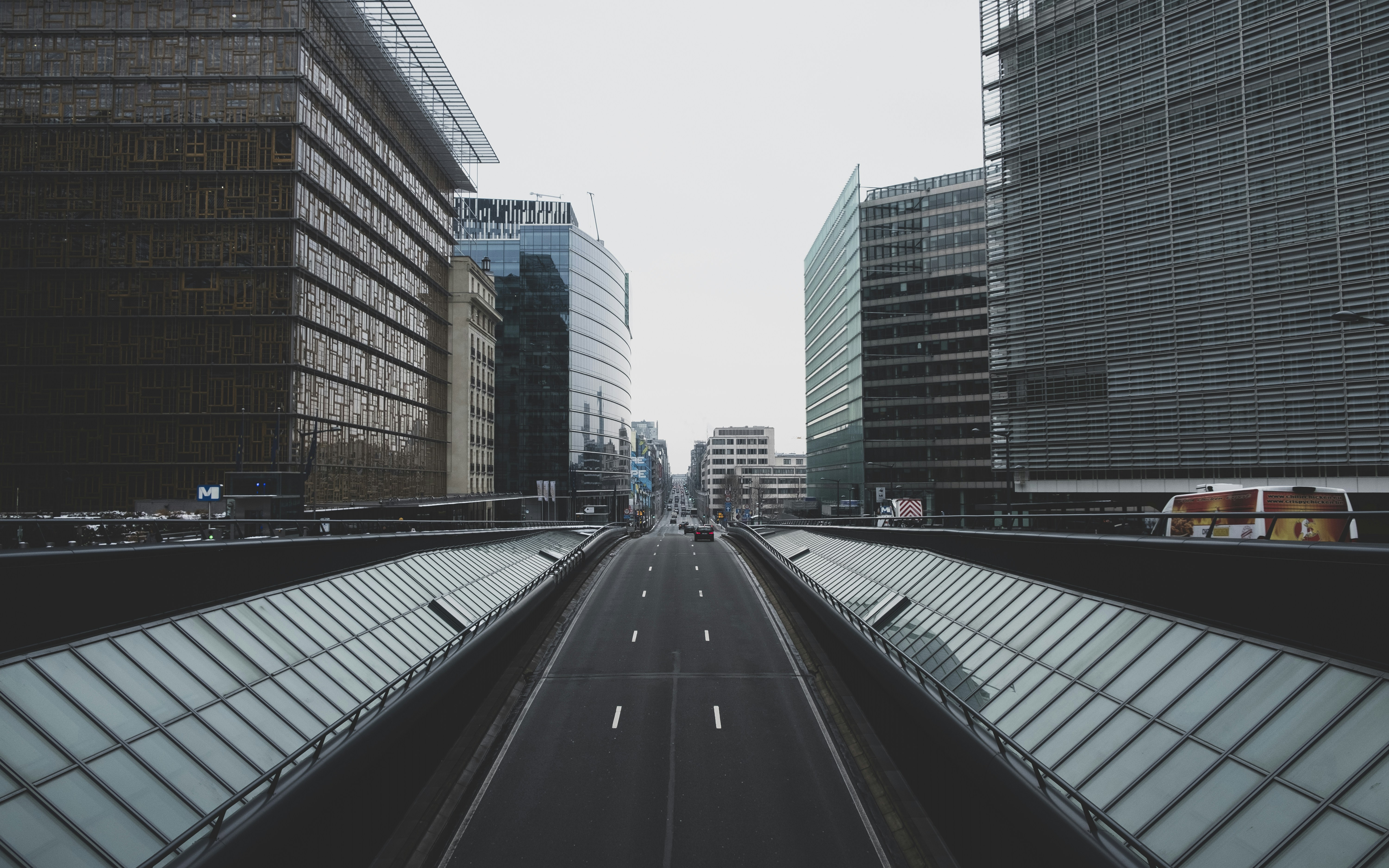 Empty city road flanked by modern glass buildings under an overcast sky.