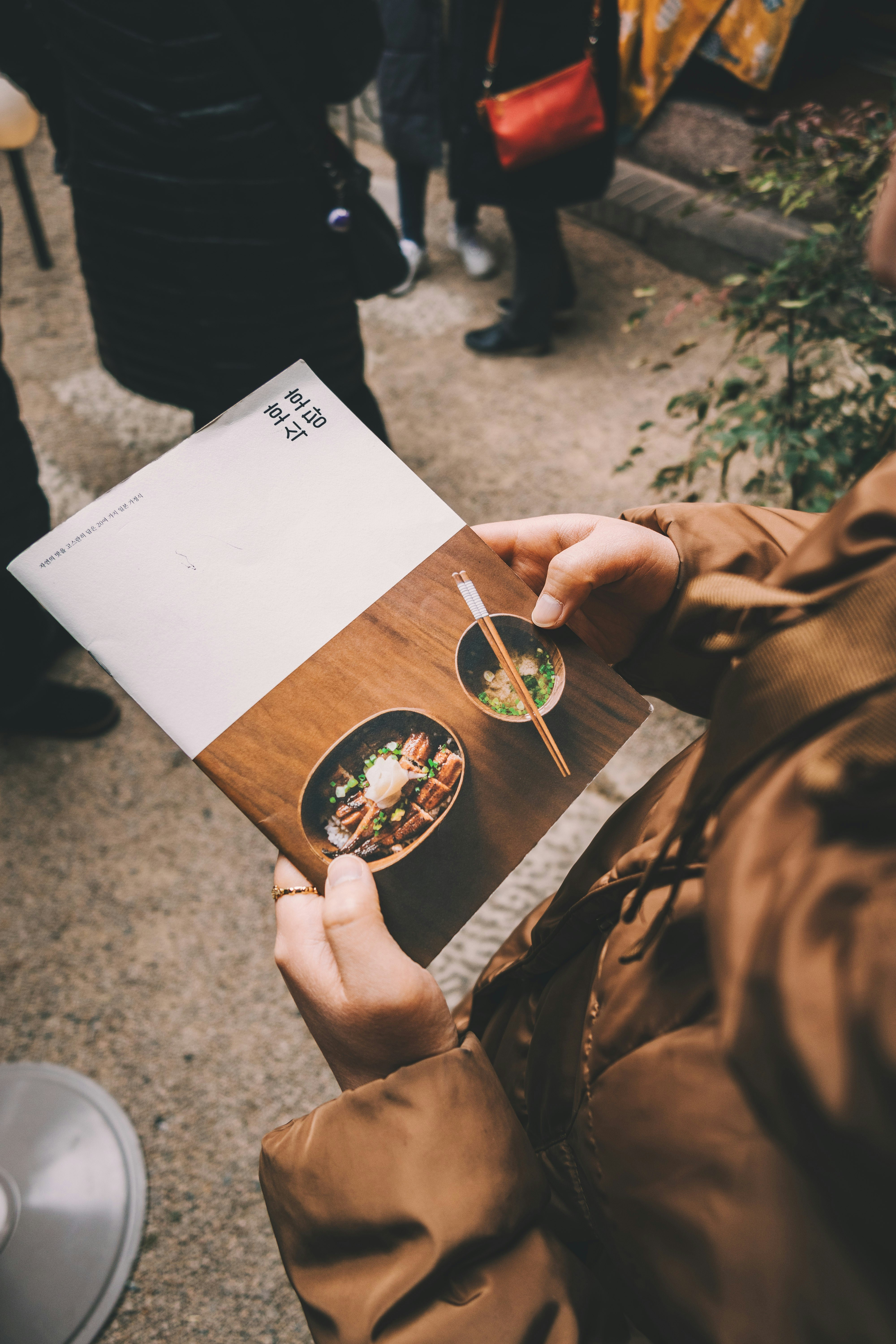 A person holding a menu featuring two bowls of vibrant dishes on a wooden table, surrounded by a bustling outdoor atmosphere.