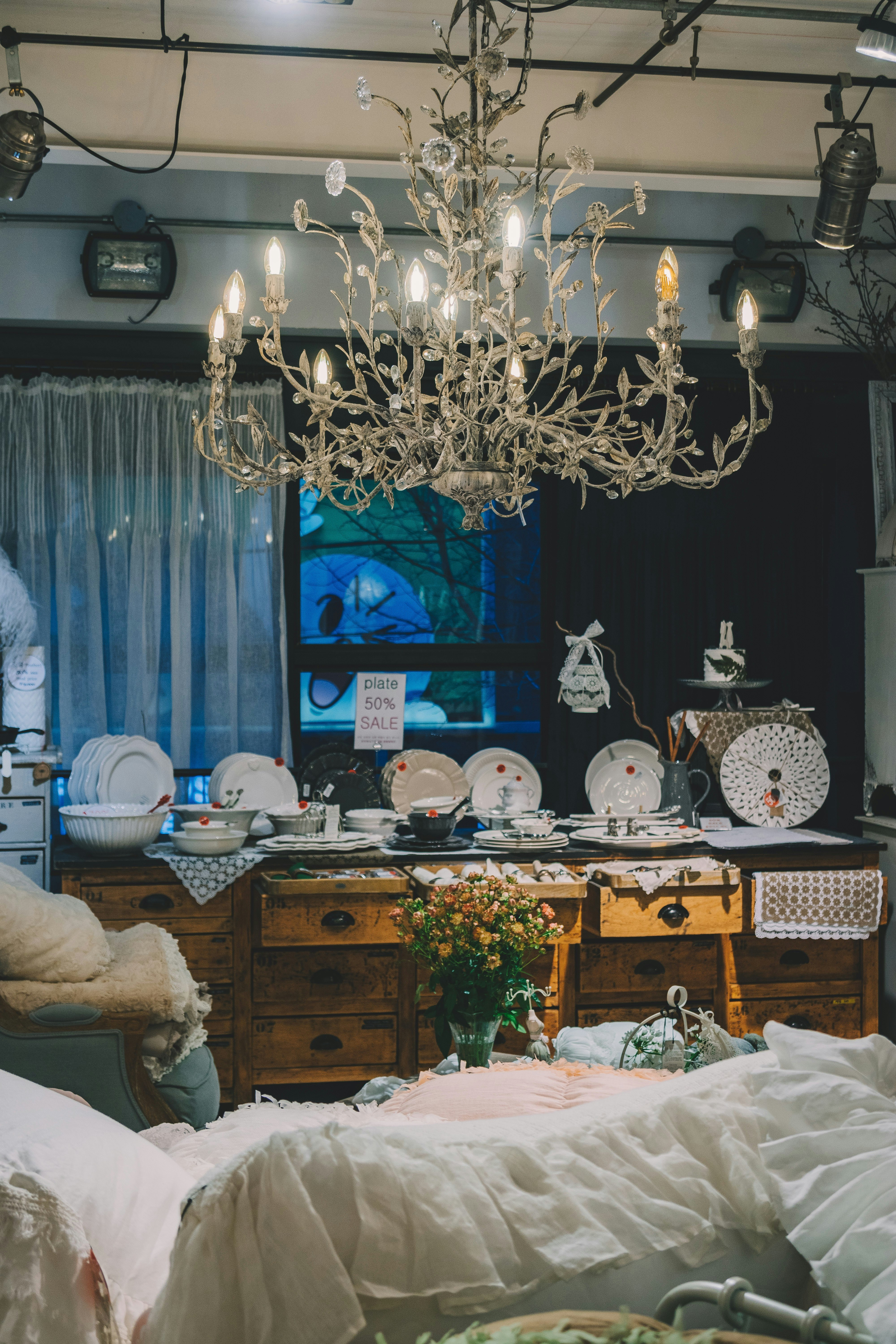 A beautifully arranged vintage shop interior featuring a decorative chandelier, wooden furniture, and an array of dishware on display. The ambiance evokes a warm, inviting atmosphere.