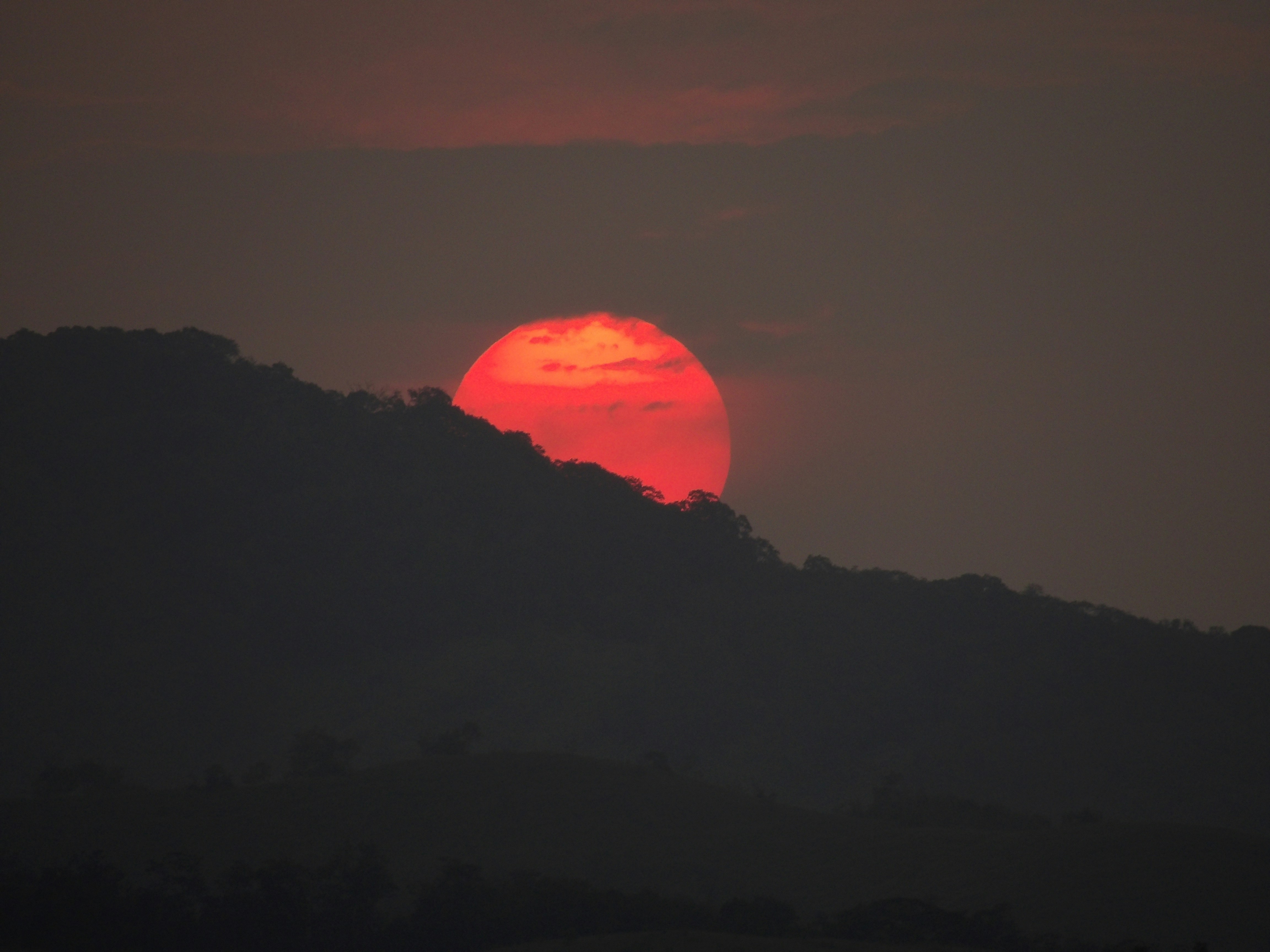 Crimson sun sinking behind silhouetted hills at dusk, with layered ridges and a hazy, warm sky.
