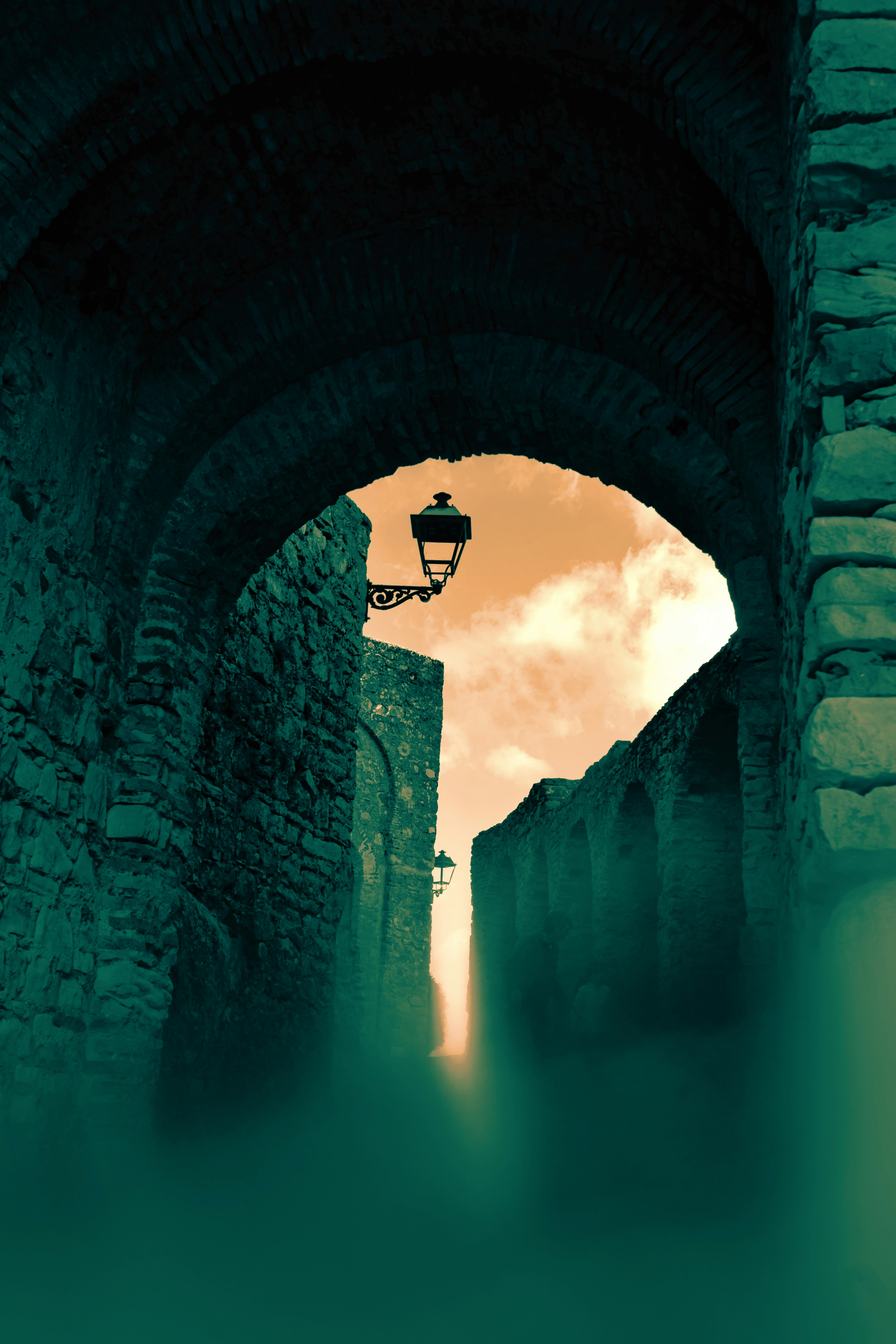 Sunlit sky peeks through a medieval archway as stone walls frame a narrow street; a lone lantern hangs above, with a teal-tinted foreground mist suggesting depth and atmosphere. A logo-like architectural scene captured in moody tones.