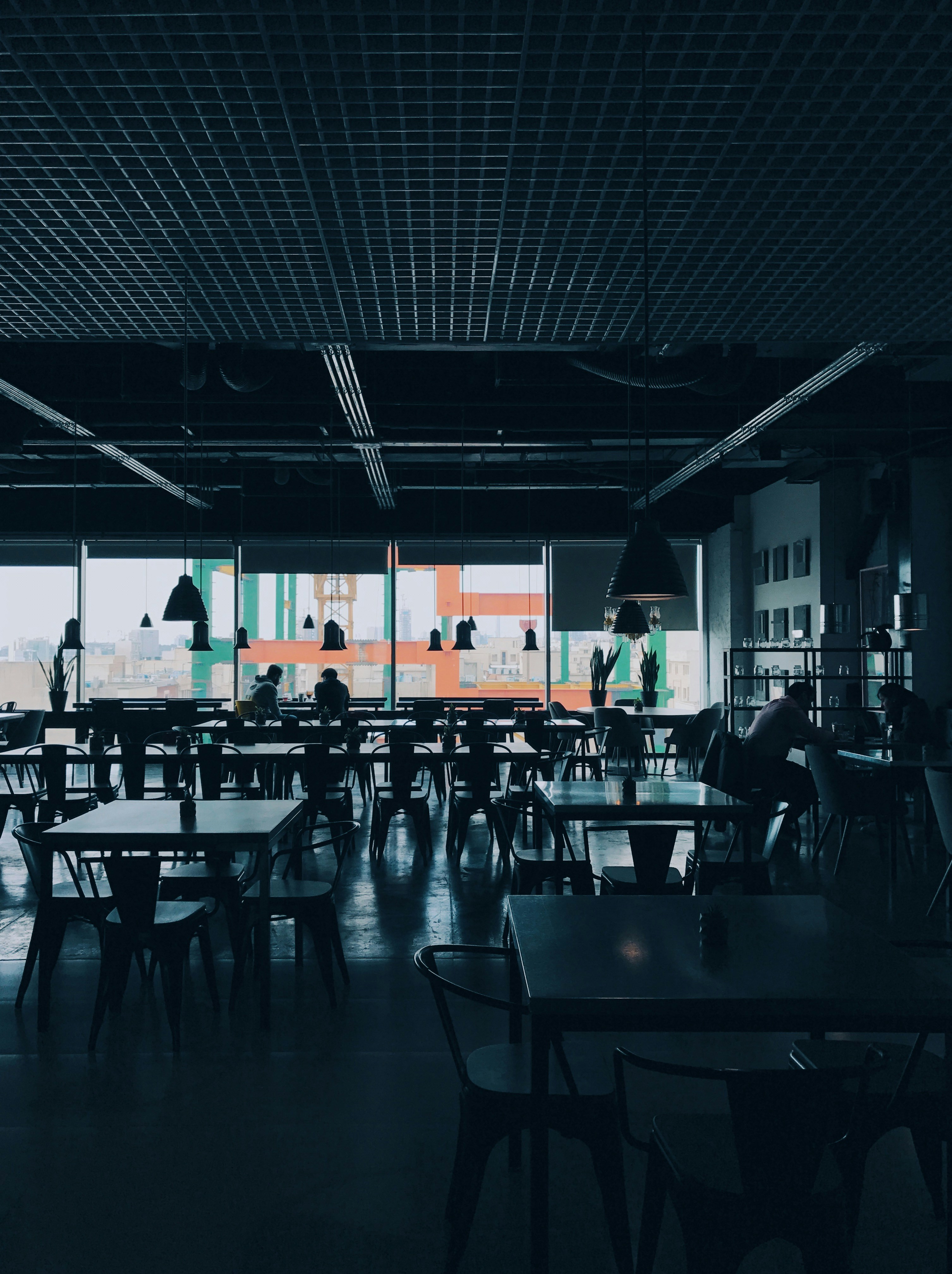 Silhouetted figures in a spacious, dimly lit café with large windows and geometric ceiling patterns.