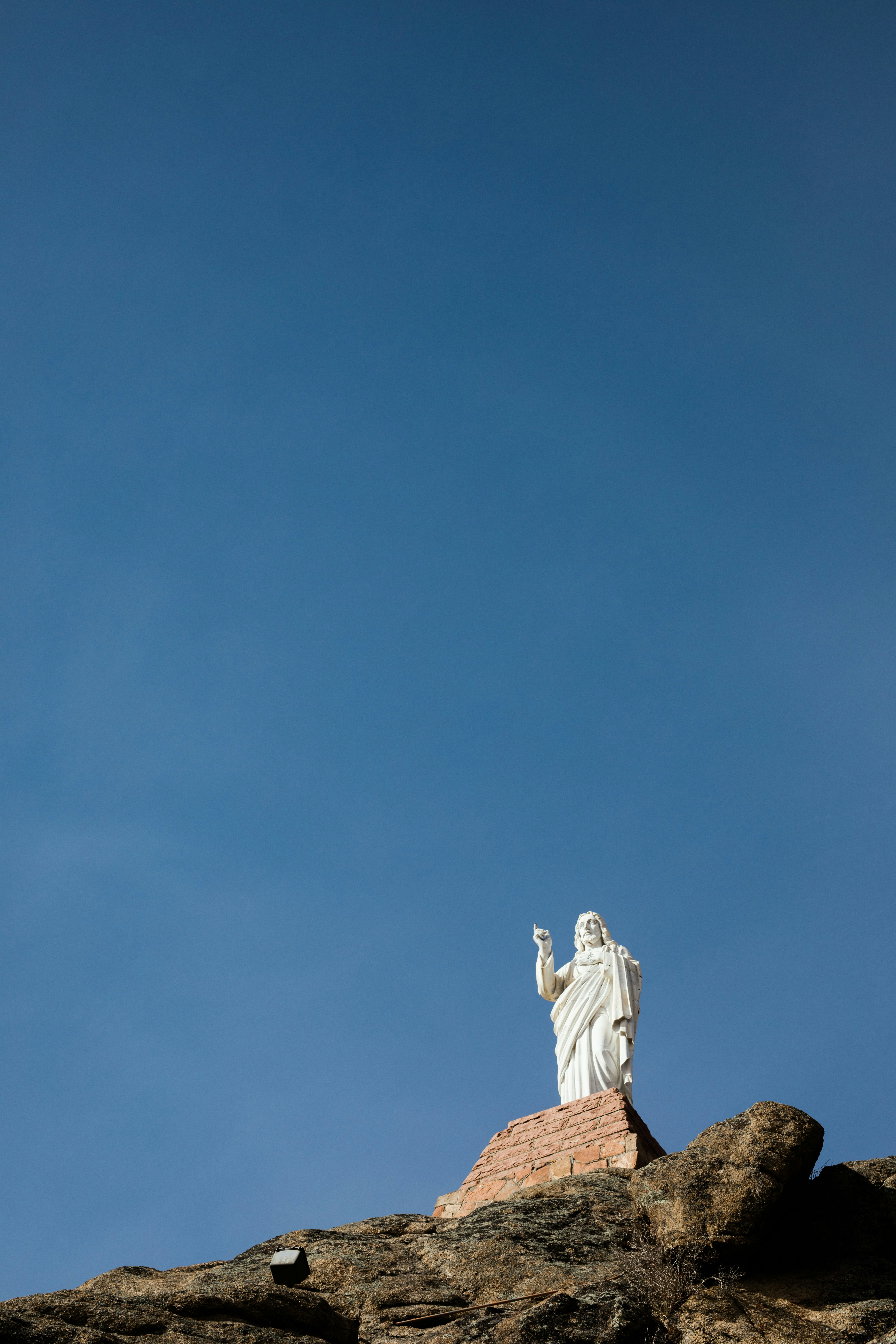 White statue of a figure with outstretched arm atop a rocky outcrop against a clear blue sky.