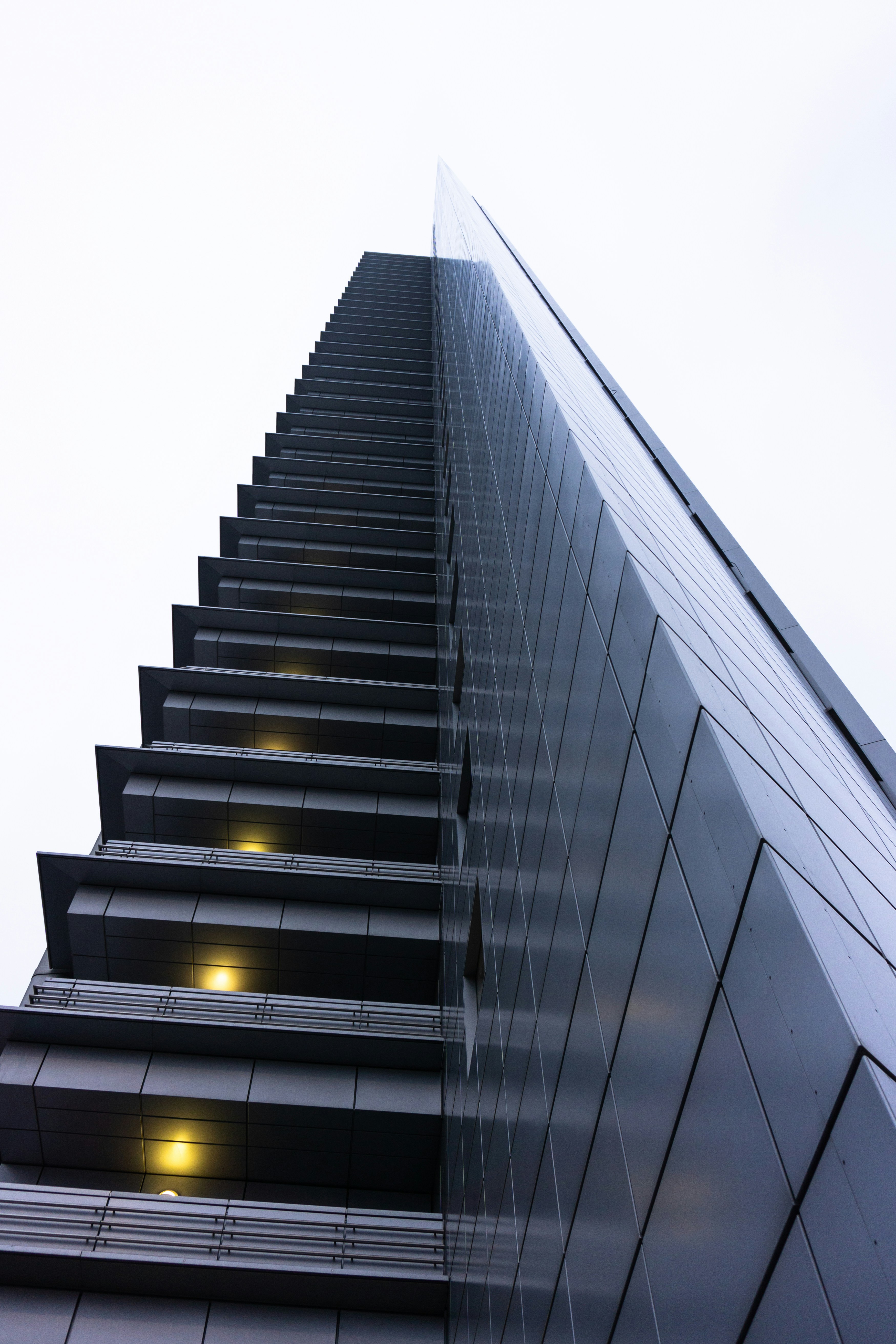 Sleek high-rise building viewed from below, showcasing its reflective glass facade and illuminated balconies against a gray sky.