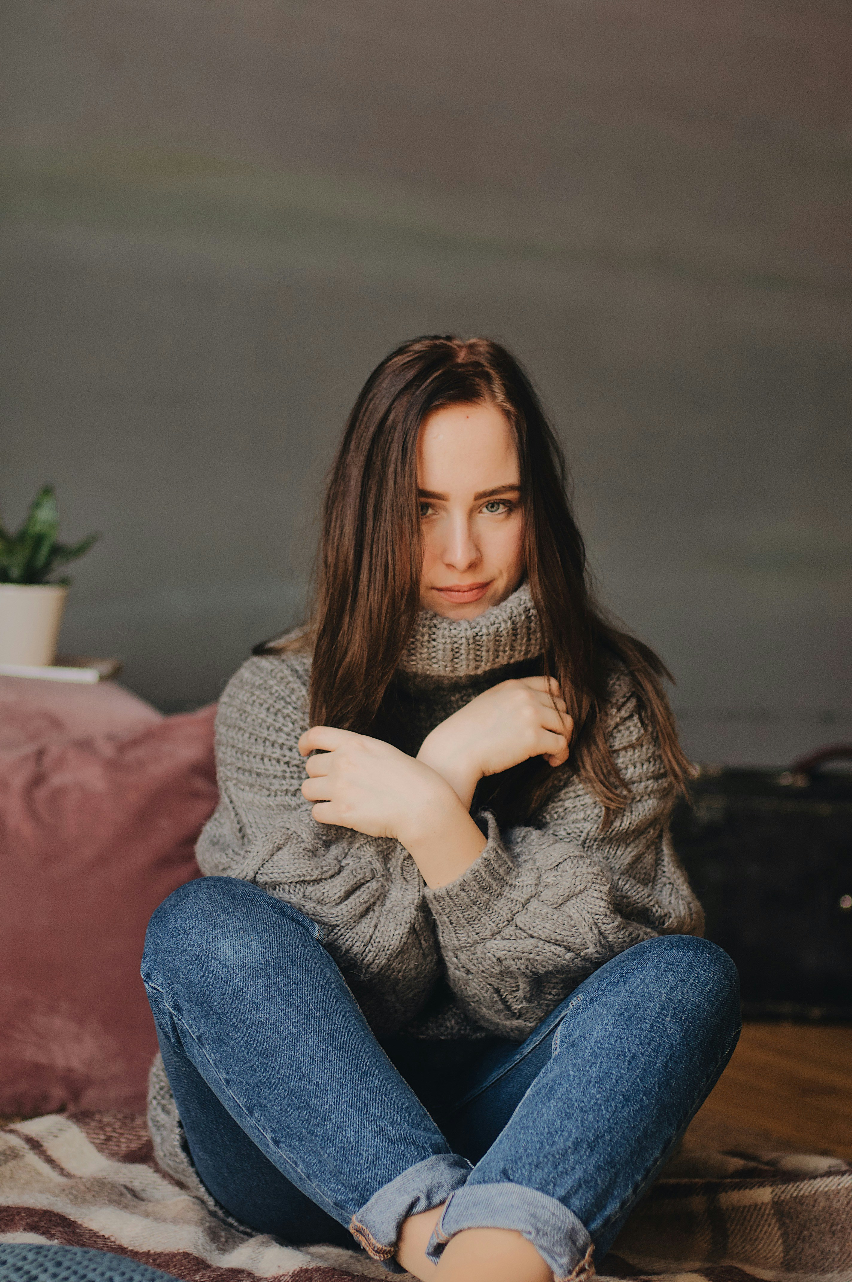 A young woman in a chunky knit sweater sits cross-legged on a blanket, exuding a sense of calm and introspection in a softly lit room.