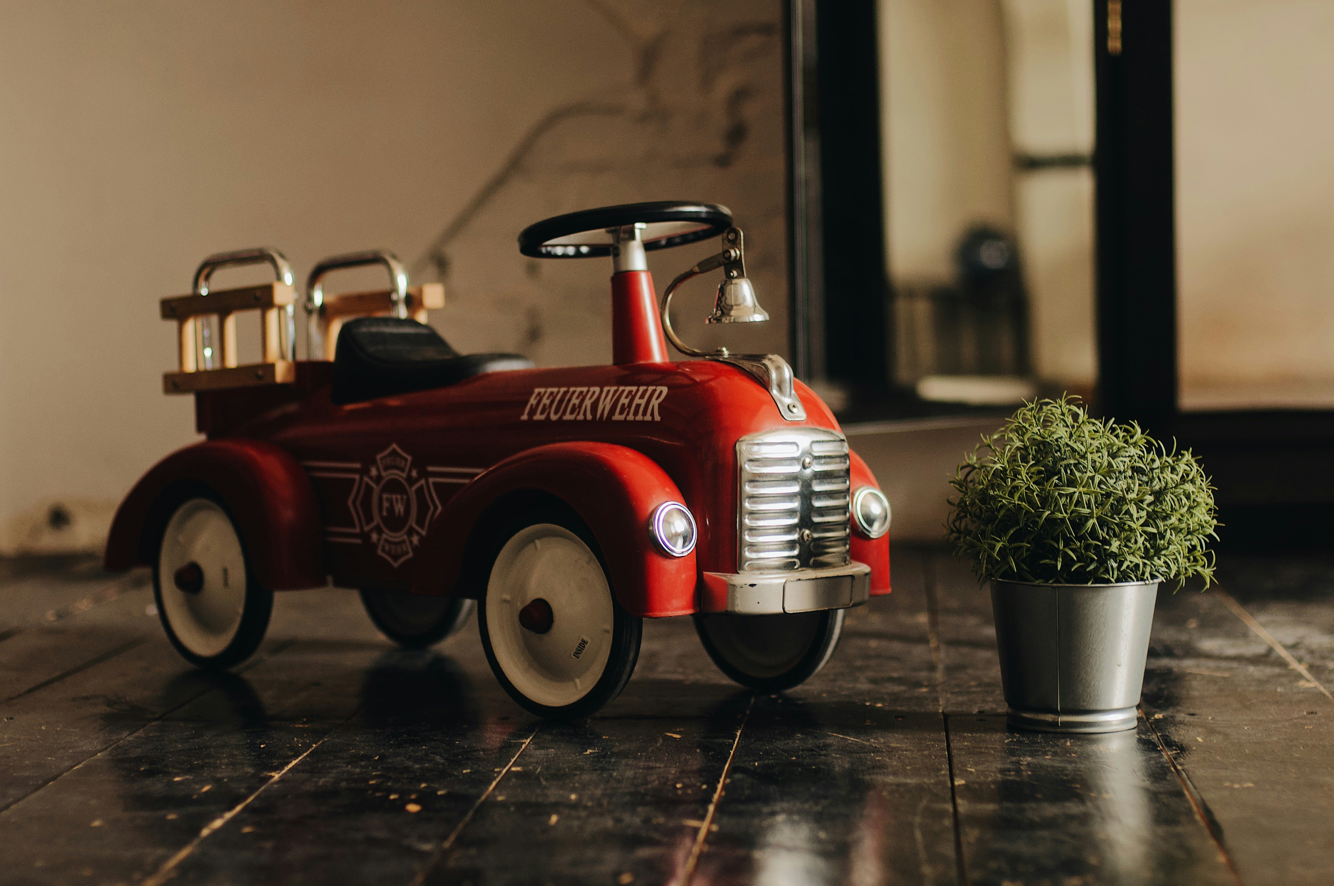 A vintage-style red firetruck toy with a shiny front grill and a small potted plant nearby, set against a wooden floor backdrop.