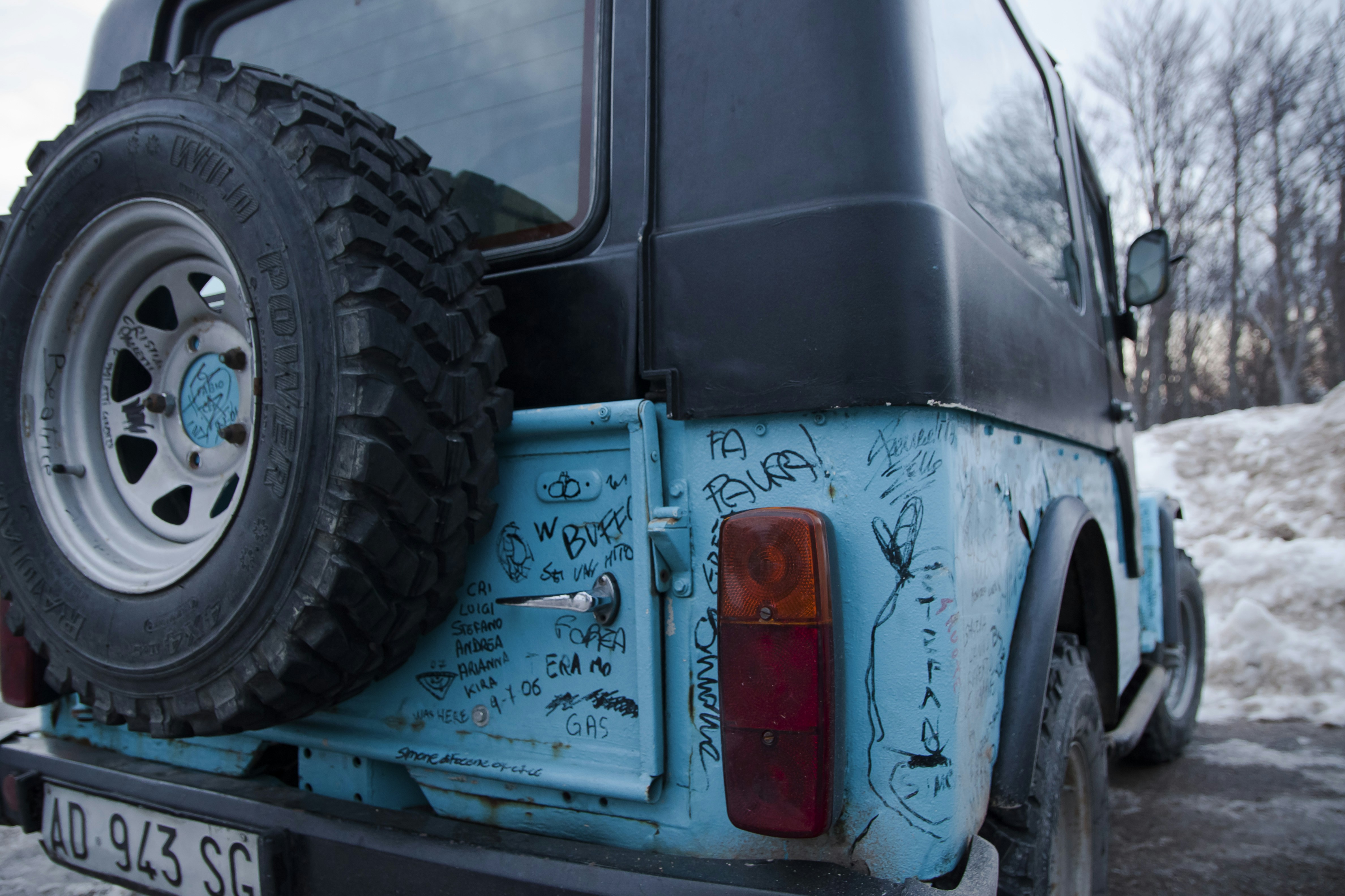 Blue off-road vehicle with graffiti parked on a snowy roadside, surrounded by bare trees.
