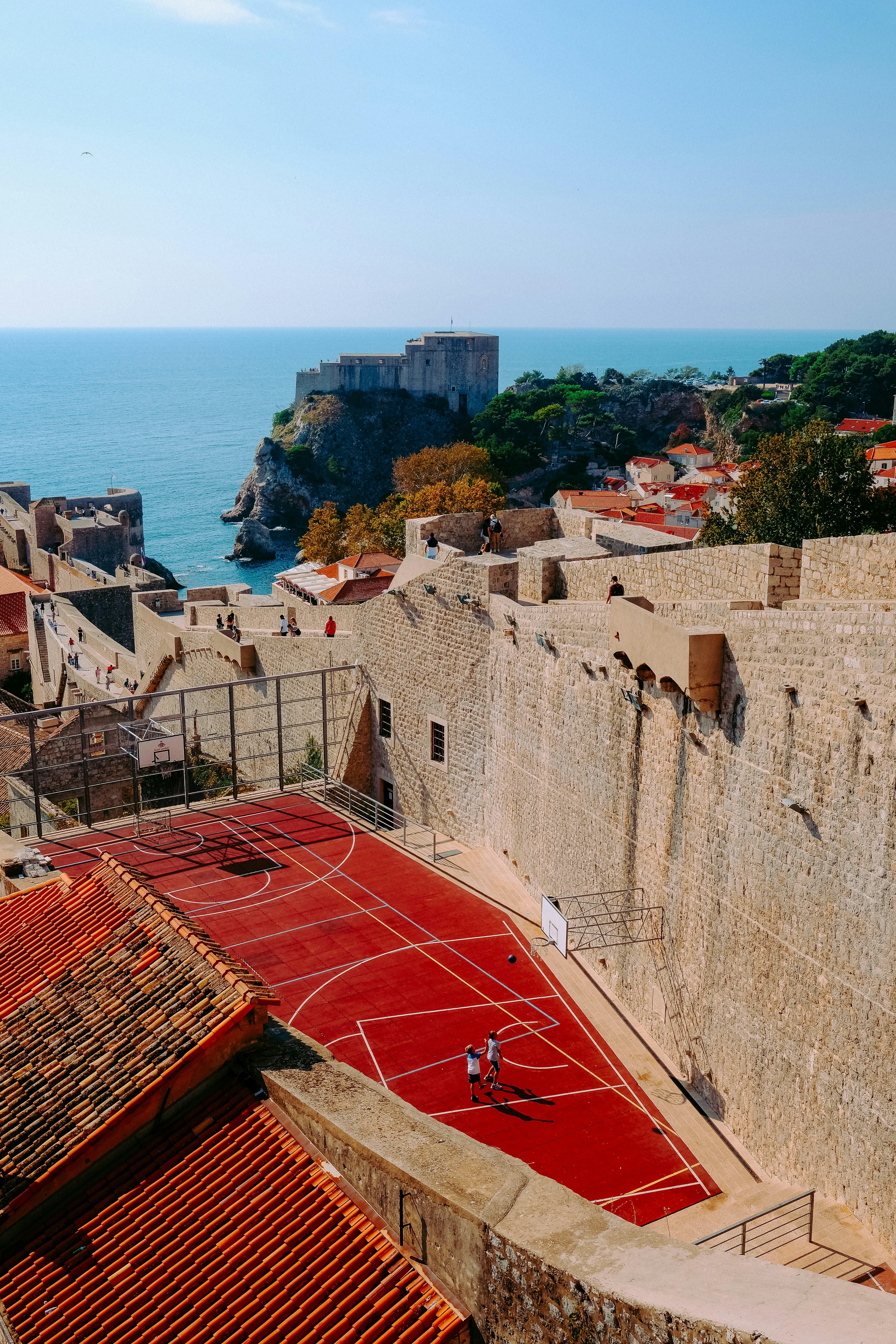 A vibrant red basketball court nestled within historic stone walls, overlooking the serene blue sea and coastal architecture.
