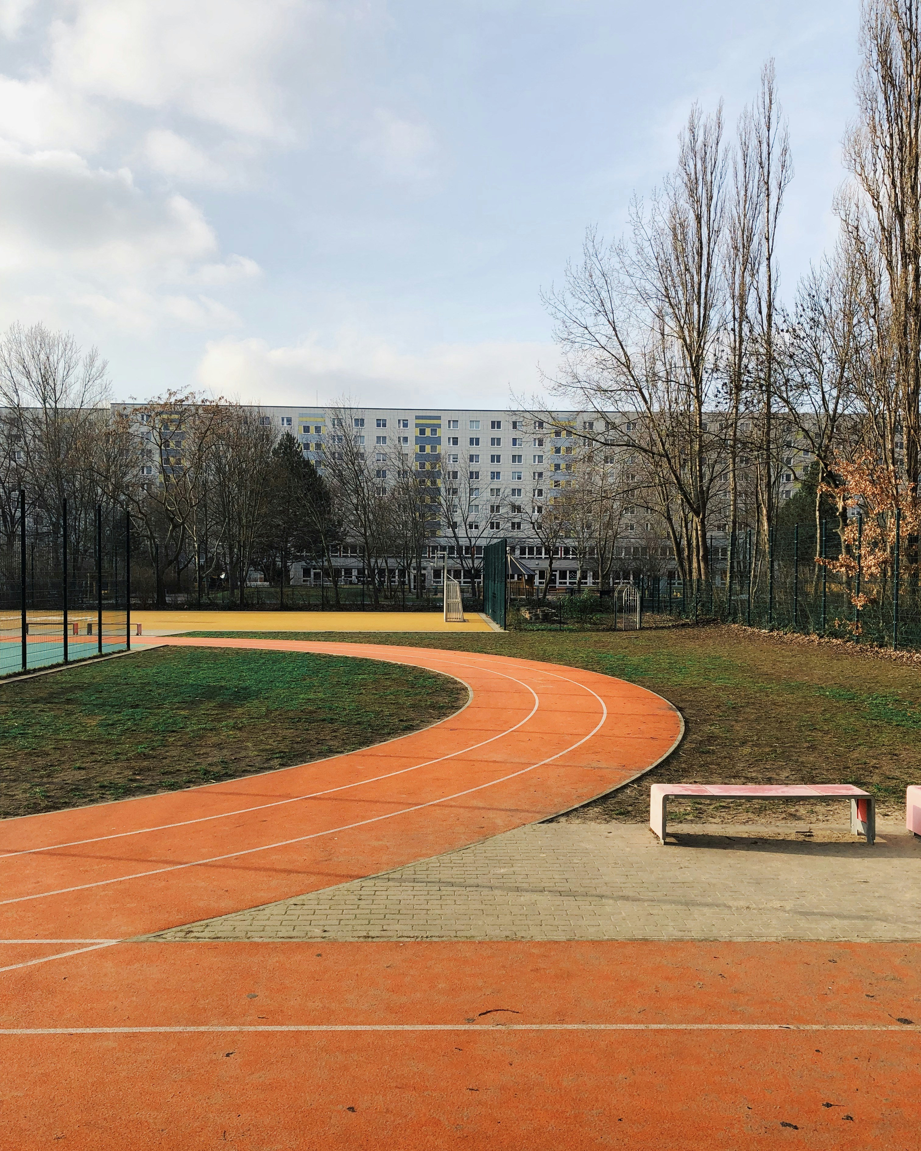 A winding orange track meanders through a community sports area, flanked by bare trees and a backdrop of residential buildings. The scene captures a tranquil moment in an urban setting.