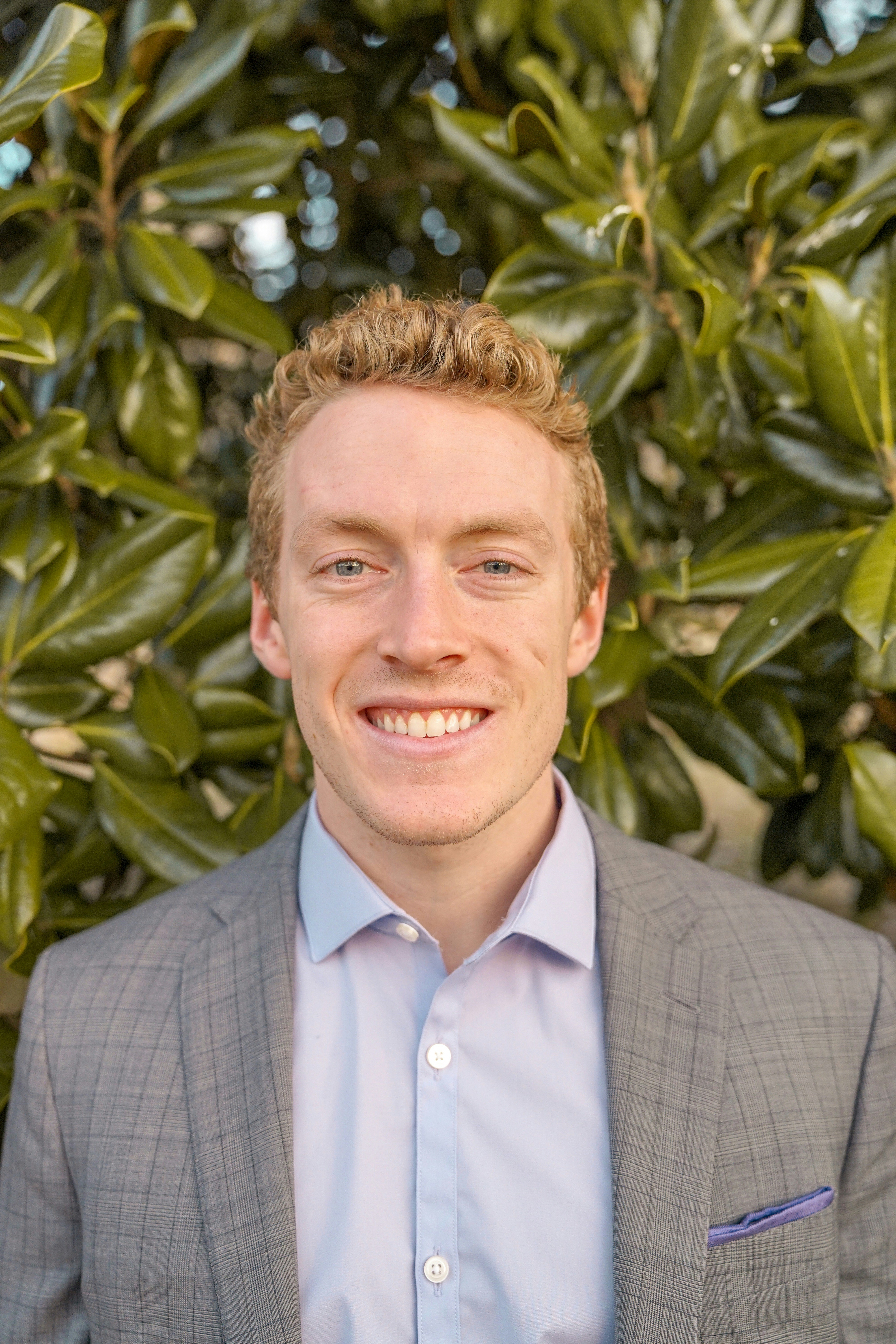 Man in a gray suit smiles confidently against a backdrop of lush green magnolia leaves.
