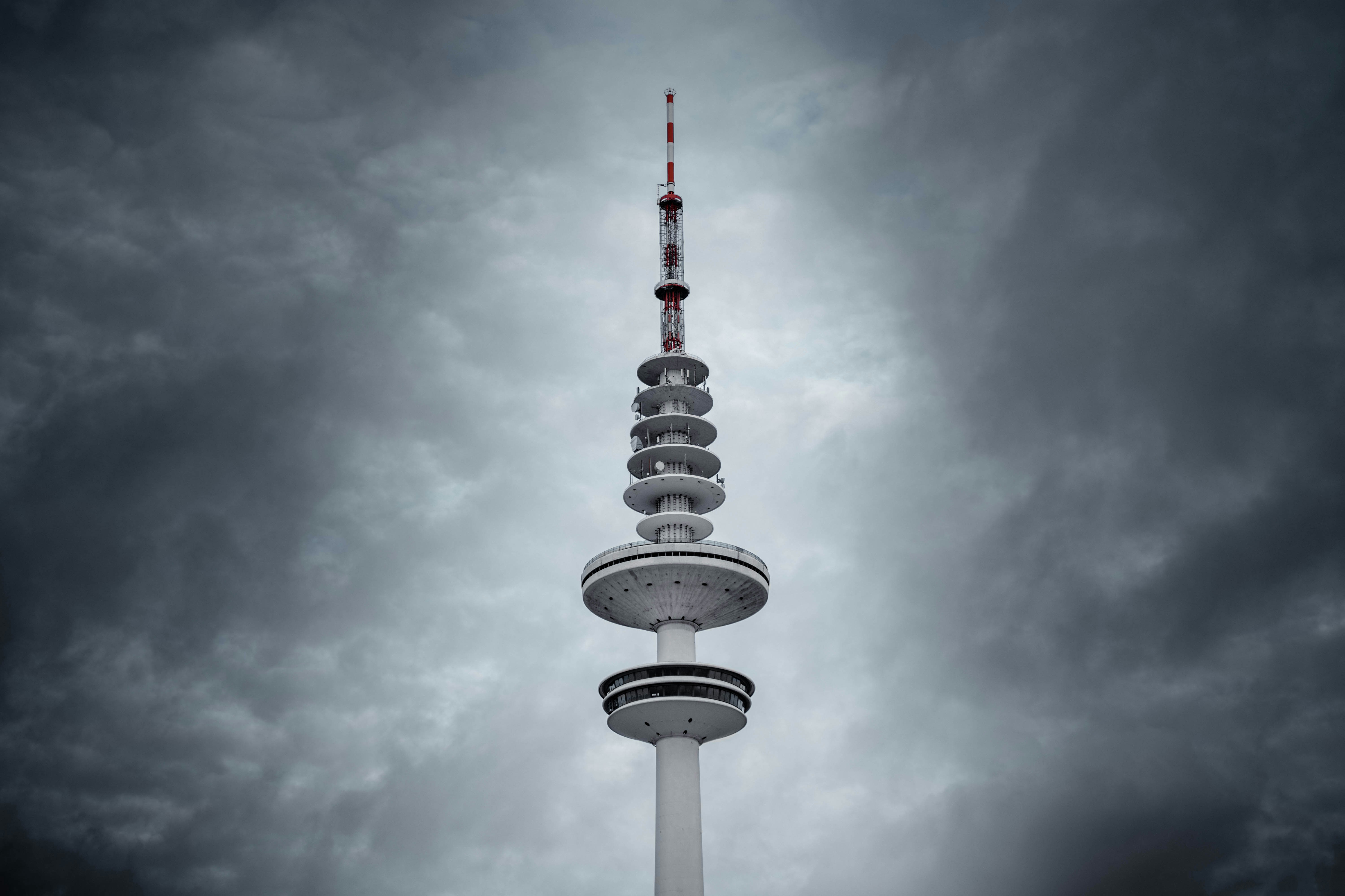 Television tower rising dramatically against a backdrop of dark, swirling clouds. The structure's design features multiple circular platforms and antennas.