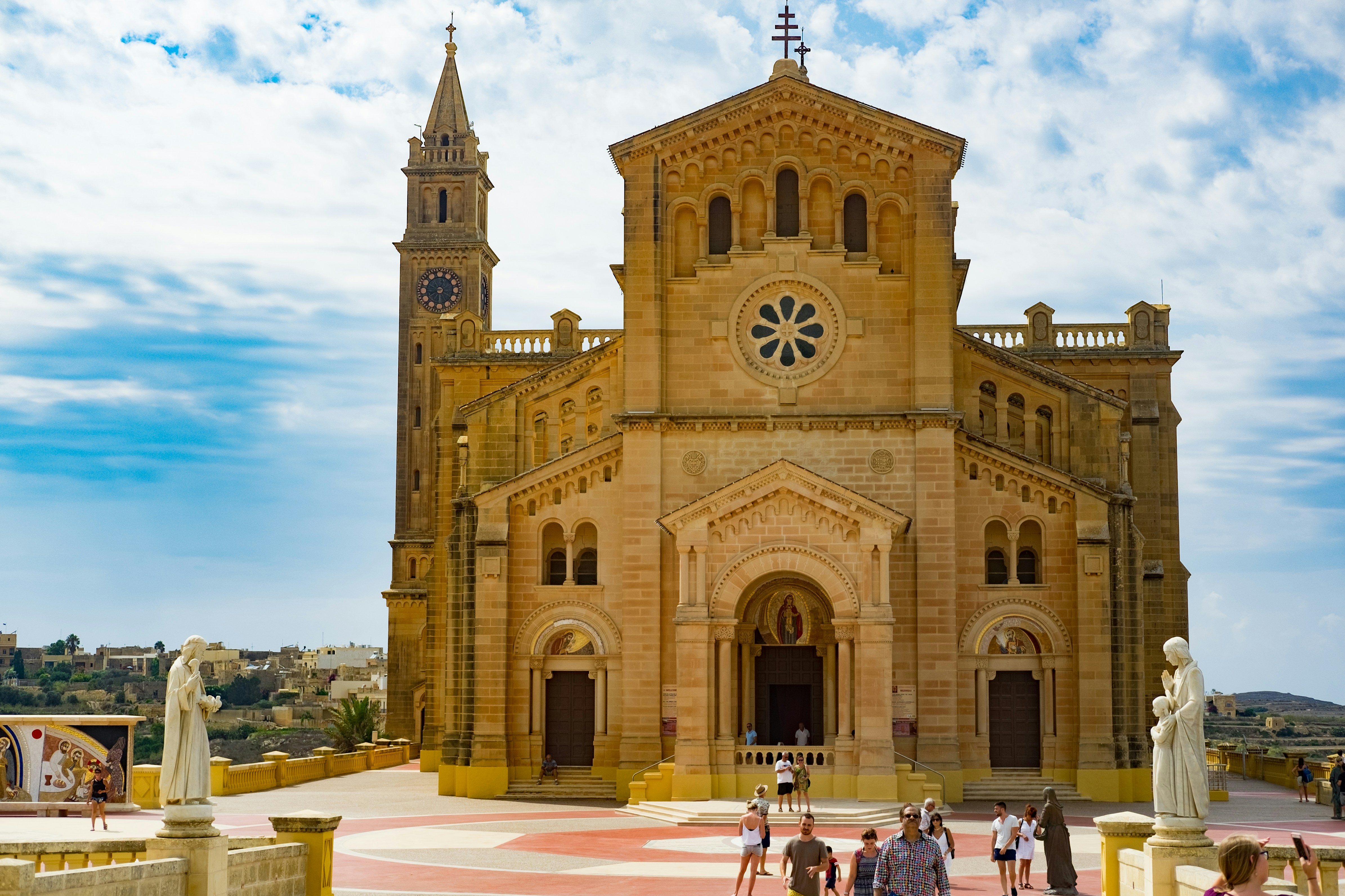 A grand church with intricate stonework and a prominent clock tower, set against a bright blue sky, filled with visitors exploring the site.