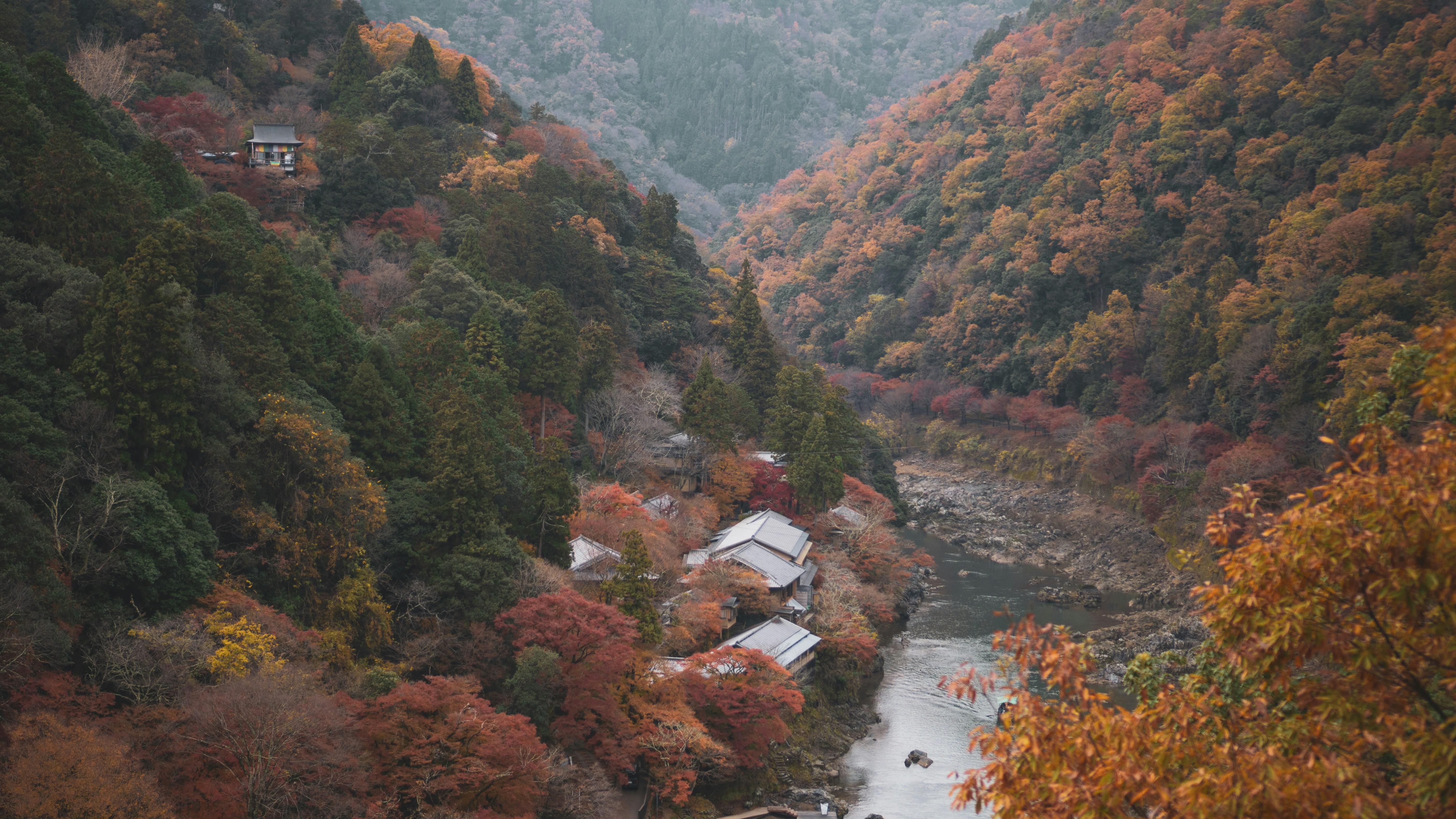 A tranquil river meanders through a vibrant valley adorned with autumn foliage, showcasing traditional structures nestled among the trees. The scene captures the essence of seasonal change.