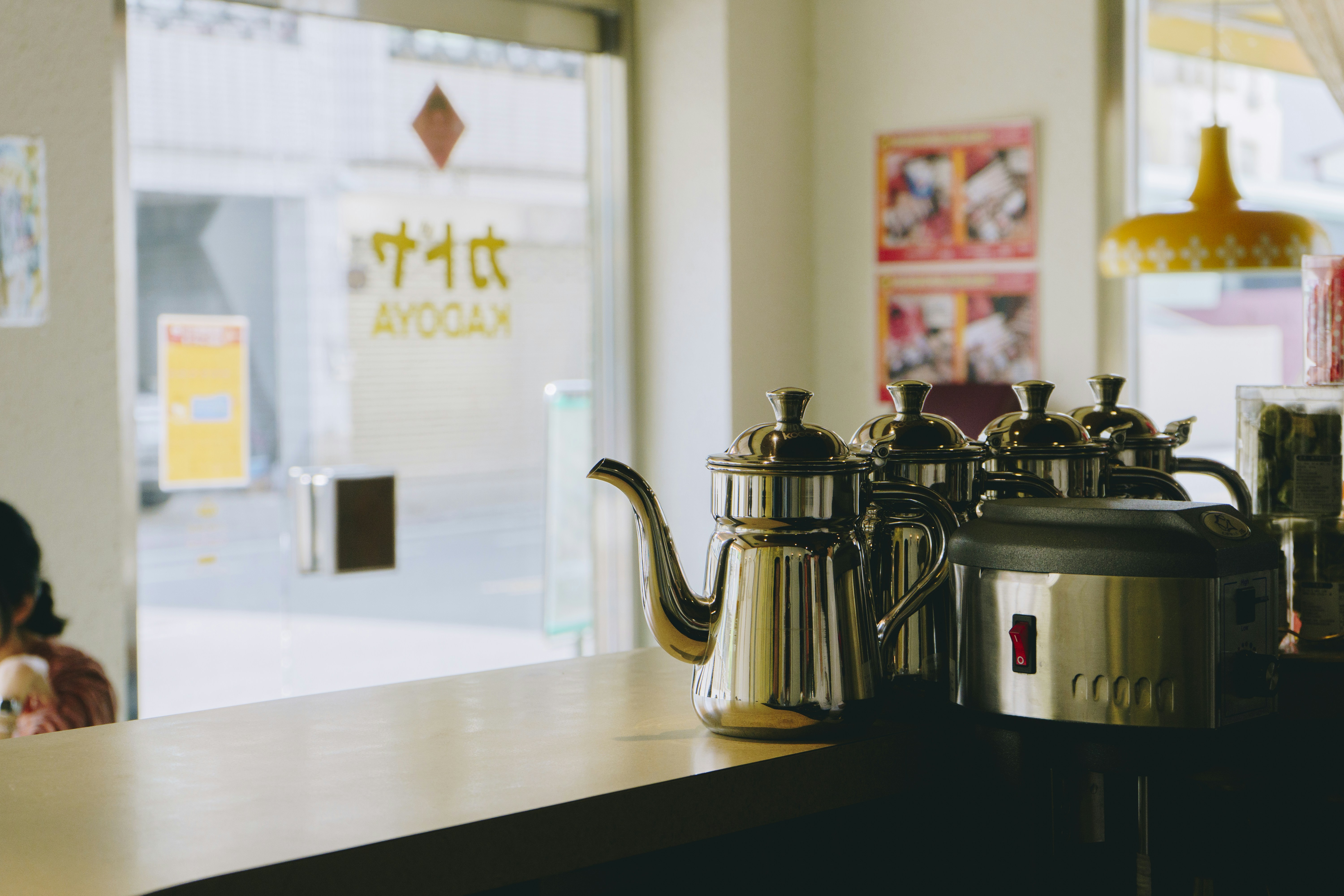 Shiny metal teapots lined up on a counter with a view of a street through a glass door. The atmosphere hints at a blend of contemporary and traditional elements.