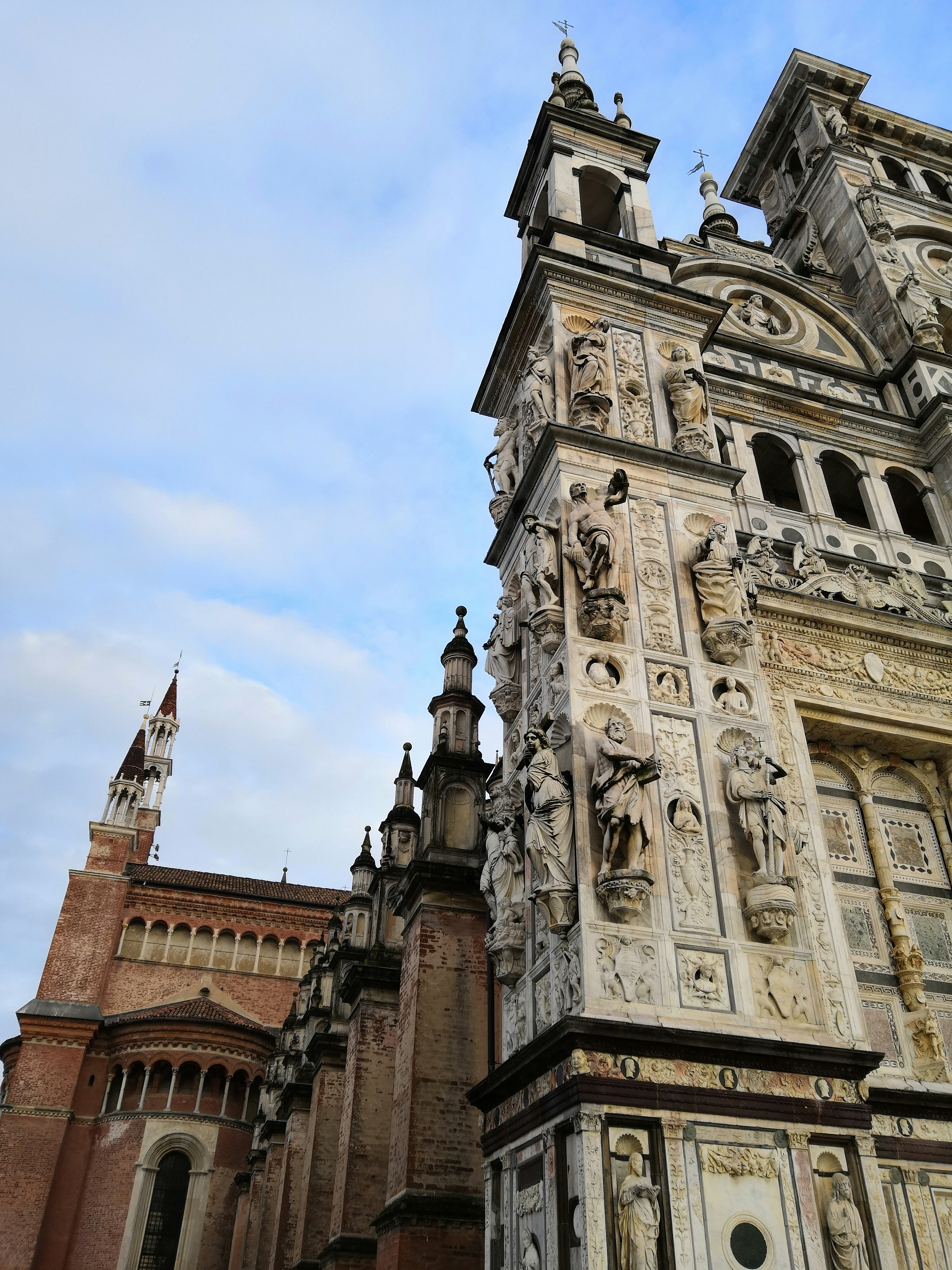A photograph of an ornate ivory marble façade with sculpted reliefs rising beside red brick Gothic wings against a bright blue sky. It highlights the depth and texture of the carvings.
