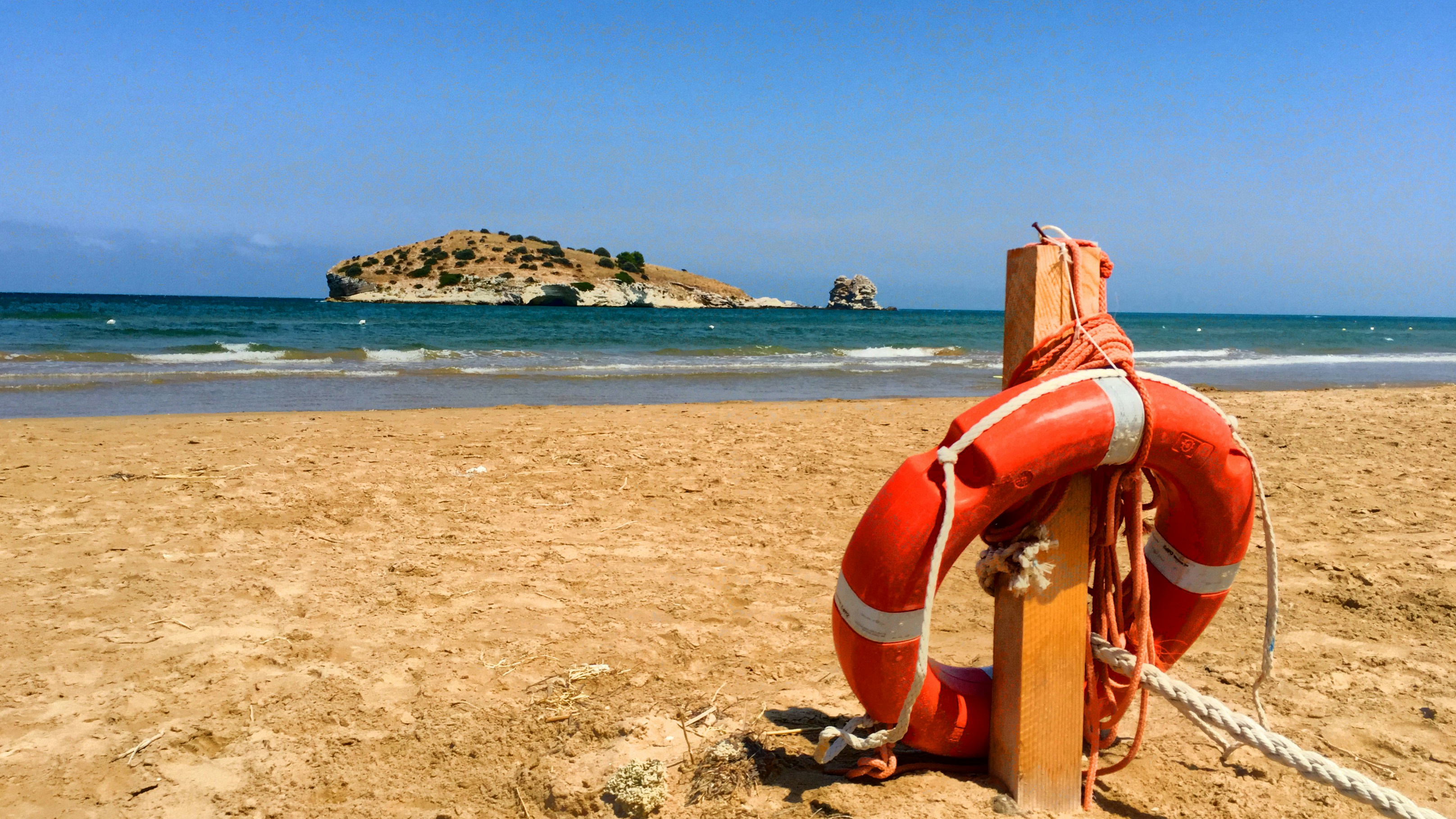 Bright orange lifebuoy mounted on a wooden post by a sandy beach with a distant island over calm blue waters.