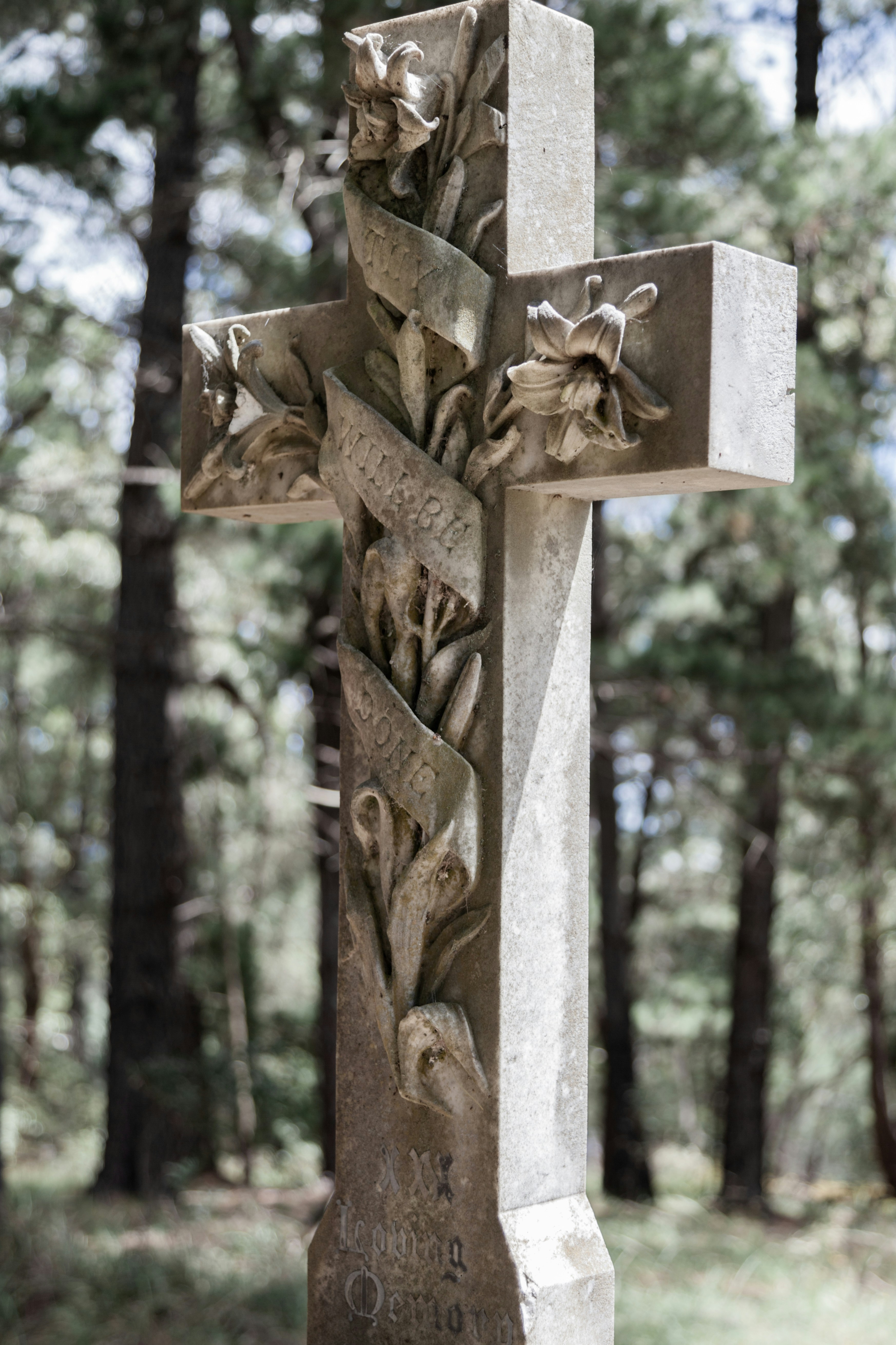 Intricately carved stone cross adorned with floral motifs in a serene forest setting. The craftsmanship reflects a timeless homage to memory.