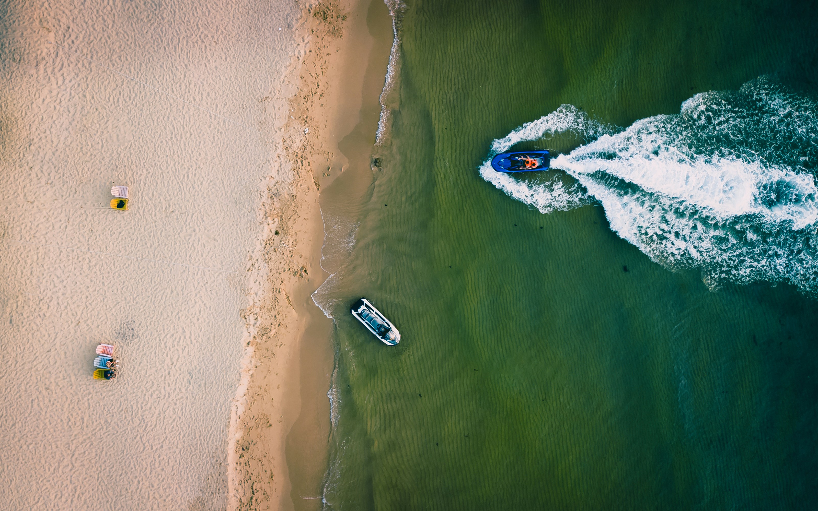 A jet ski speeds through emerald waters near a sandy beach, with a small boat anchored nearby. Colorful beach umbrellas dot the shoreline.