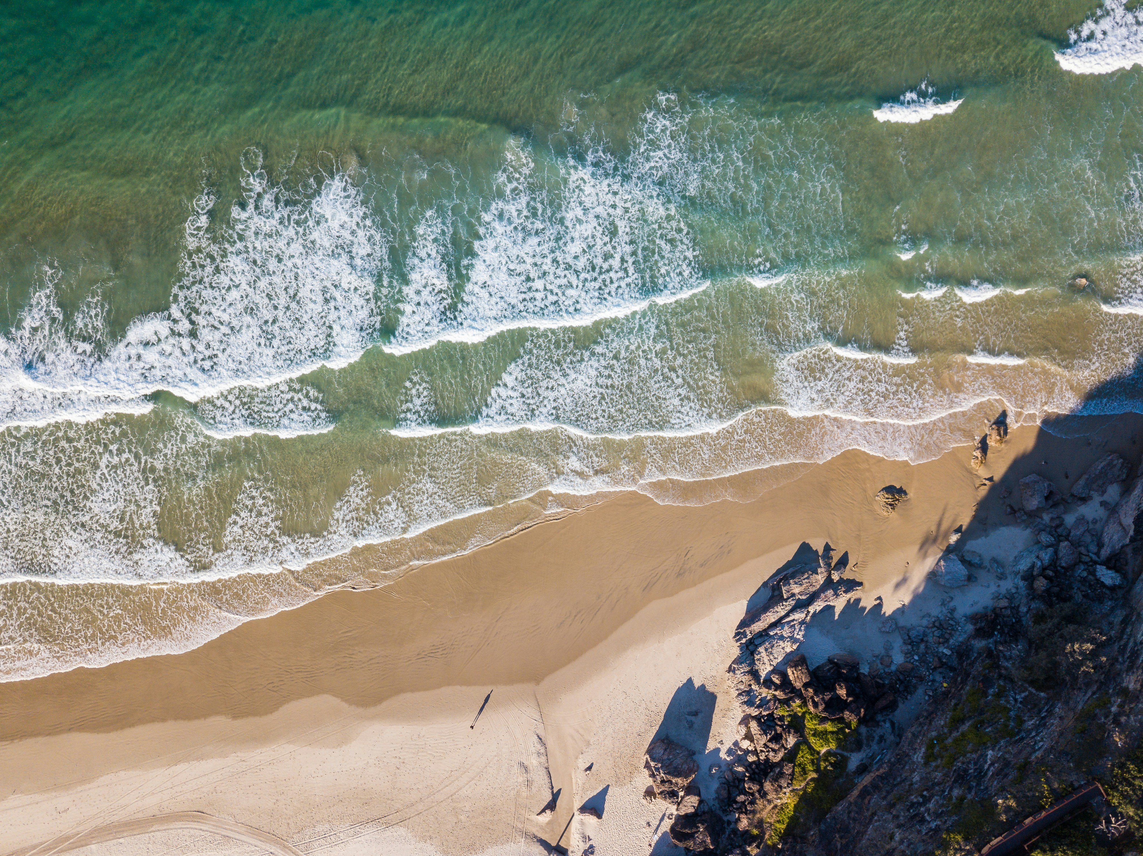 Waves gently lap against a sandy beach, revealing a rocky outcrop and coastal vegetation. The aerial perspective captures the dynamic interplay between land and sea.