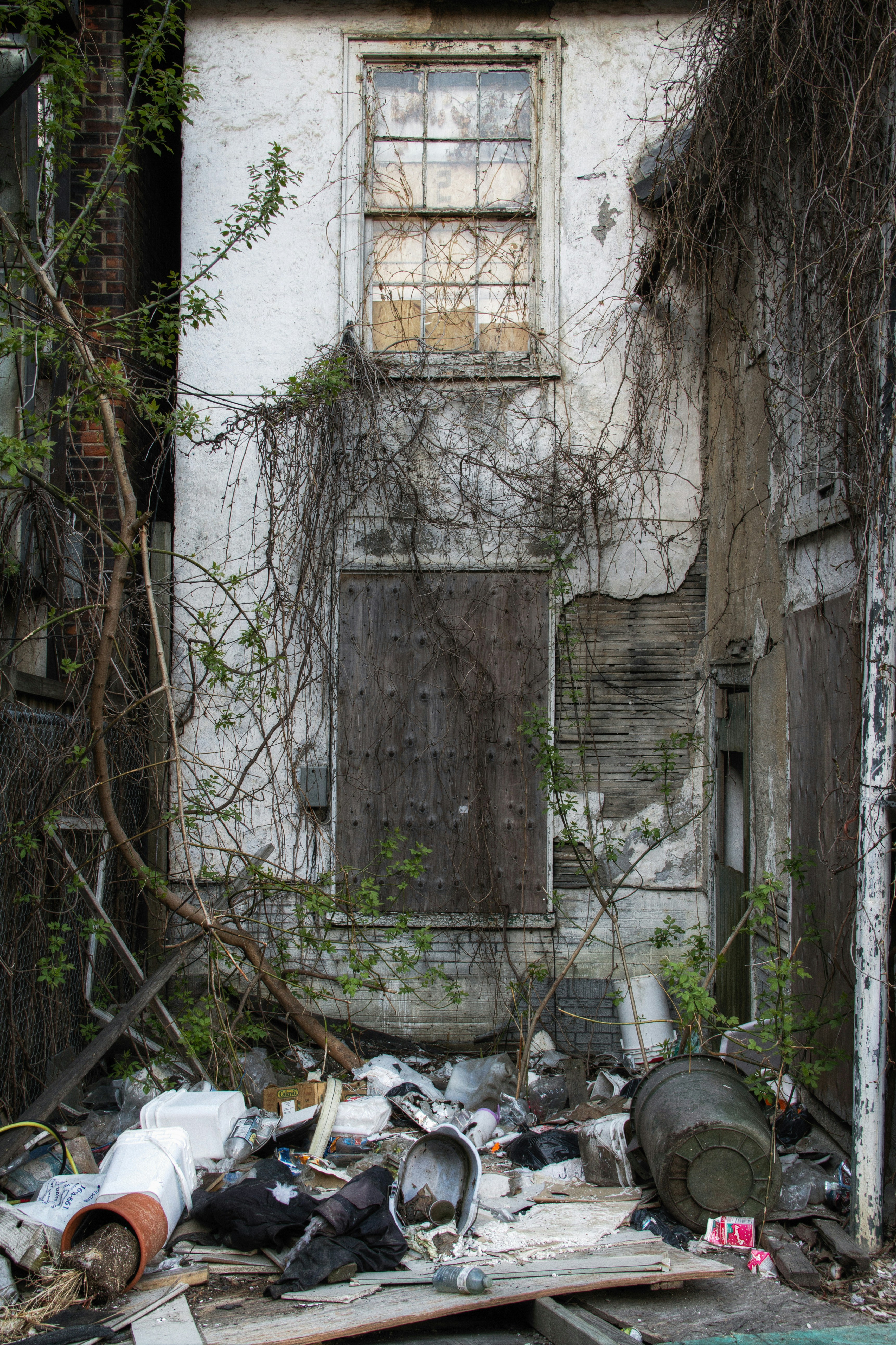 Dilapidated building with a boarded window, overrun by vines and surrounded by debris, reflecting urban decay.