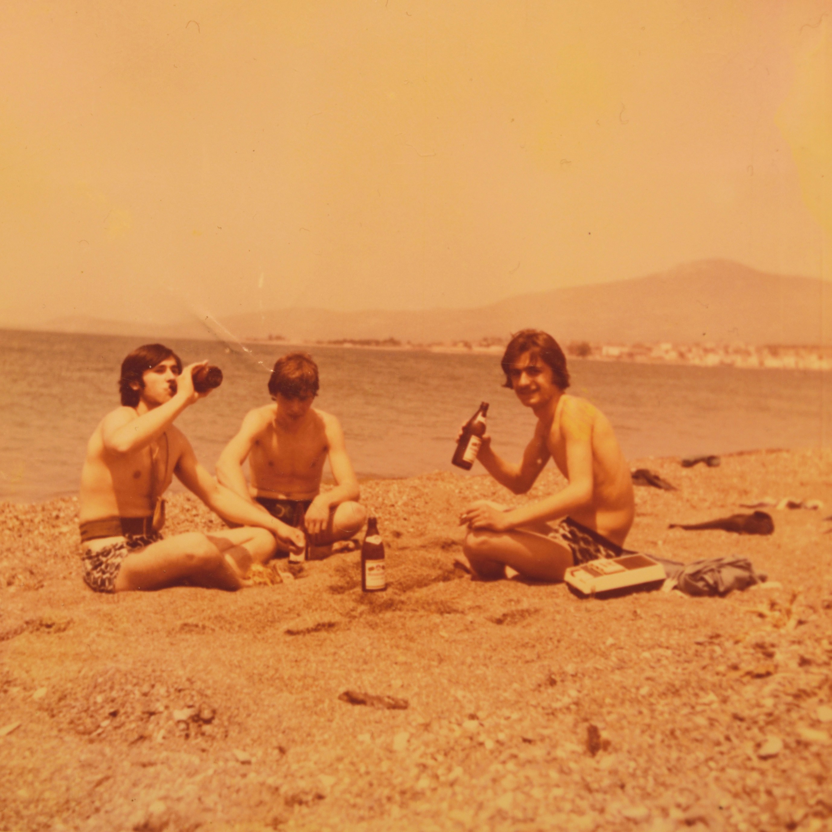 Three friends enjoying drinks on a sandy beach, basking in the sun with the ocean in the background.
