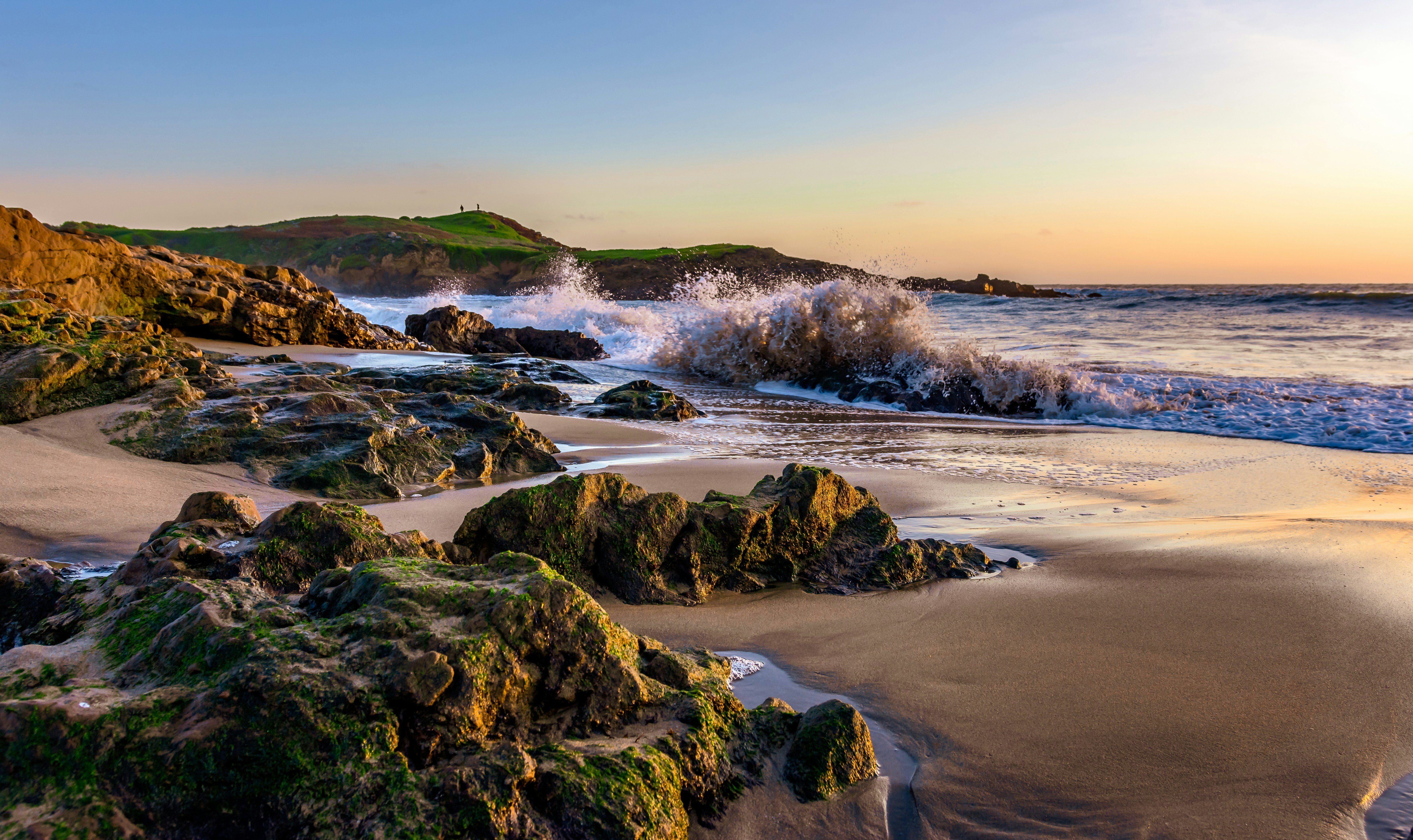 Ocean waves crashing against moss-covered rocks on a golden sandy beach at sunset.