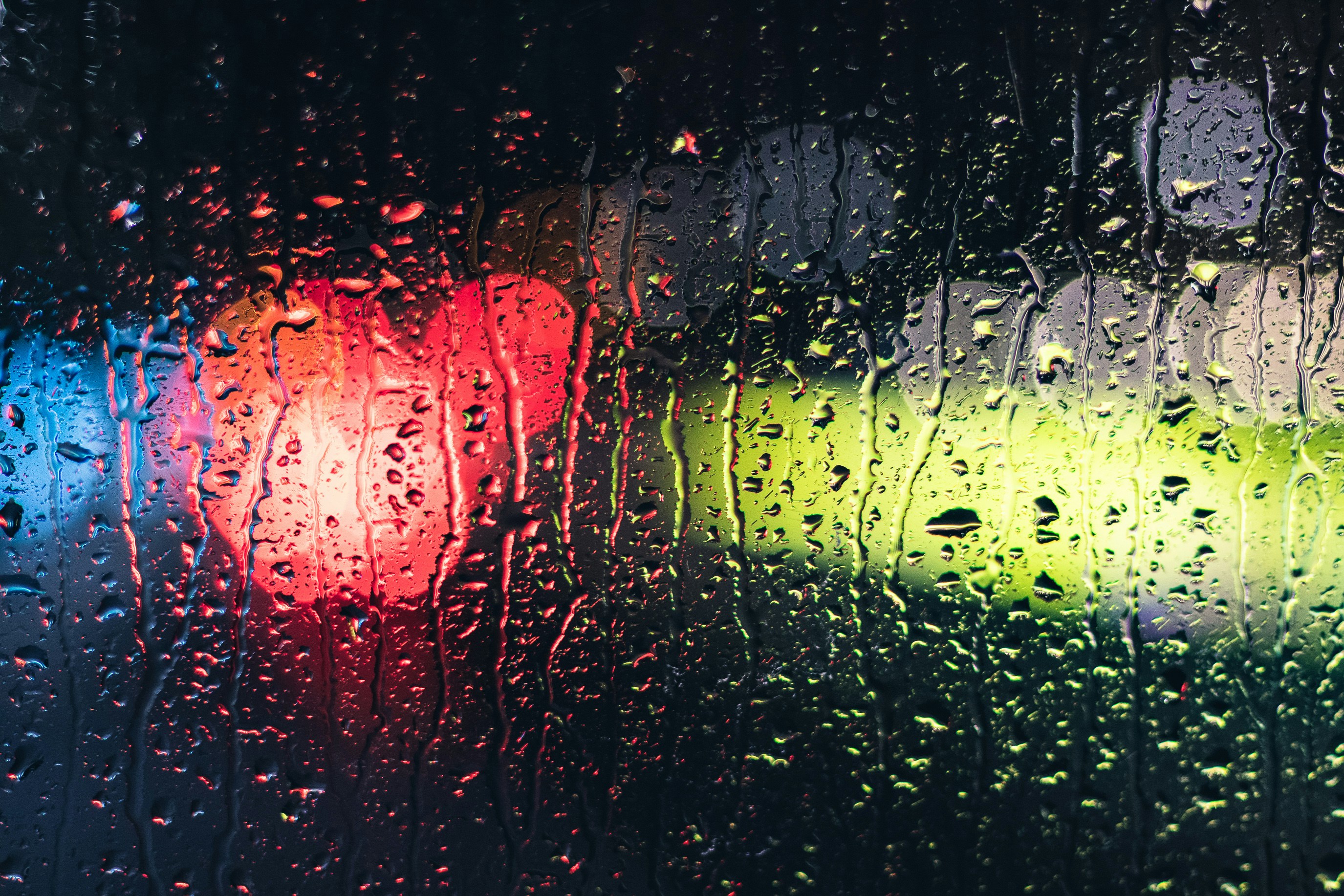 Colorful lights blurred by raindrops on a window, creating an abstract, atmospheric scene. The interplay of light and water adds depth and mood.