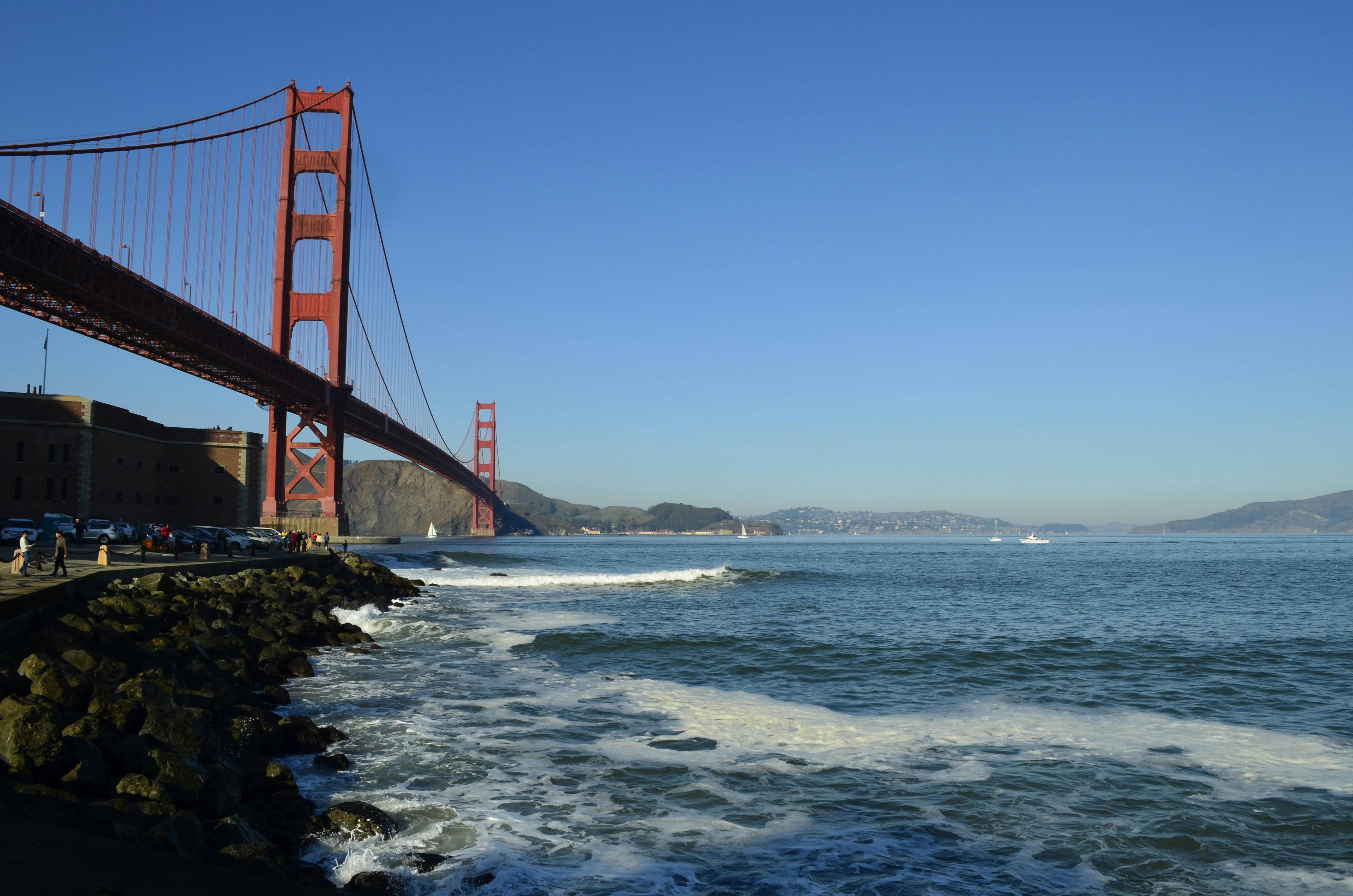 Golden Gate Bridge towering against a clear blue sky, with waves lapping at the rocky shore below.