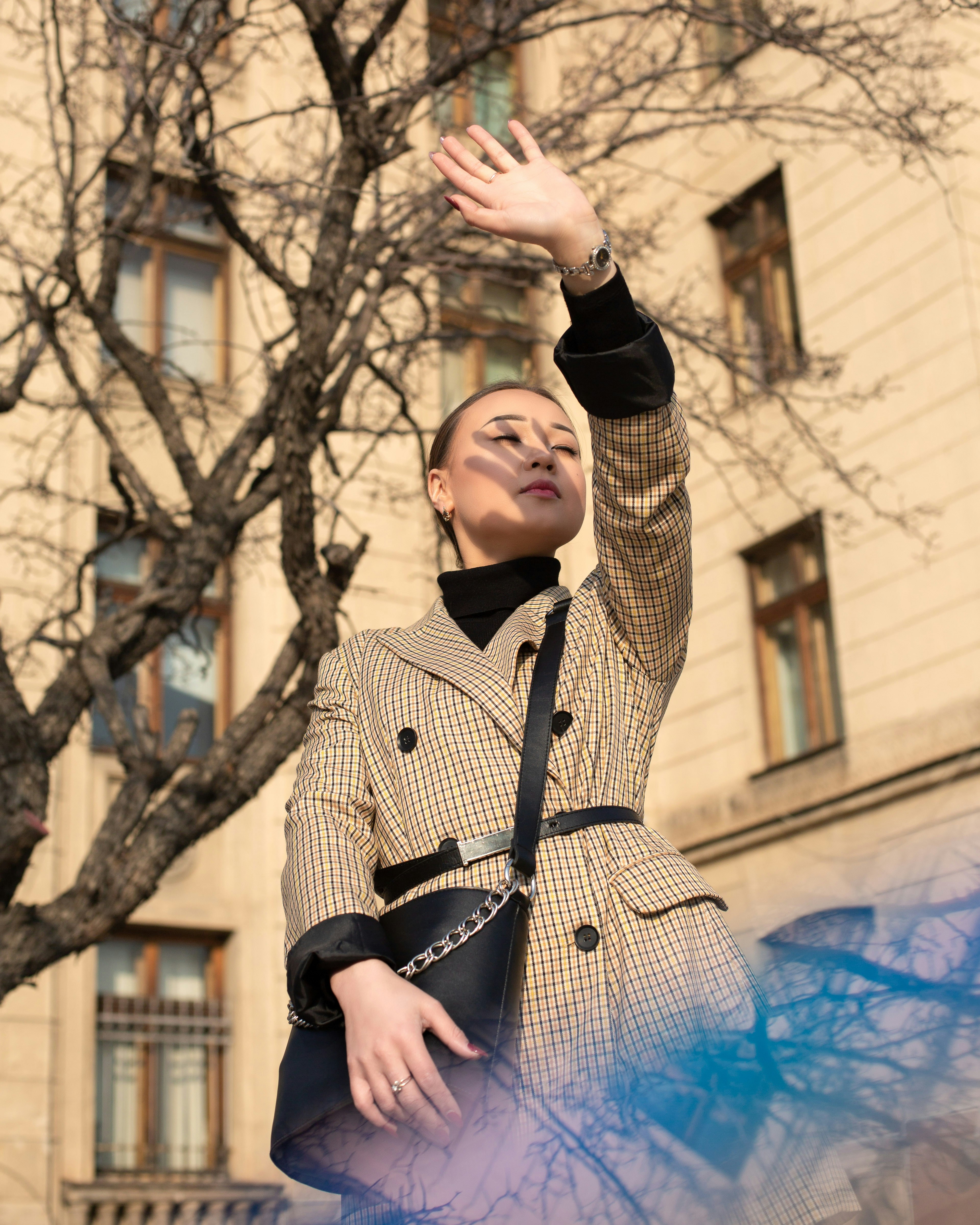 Fashionable woman in a stylish plaid coat poses against a historic building, hand raised to shield her eyes from the sun.