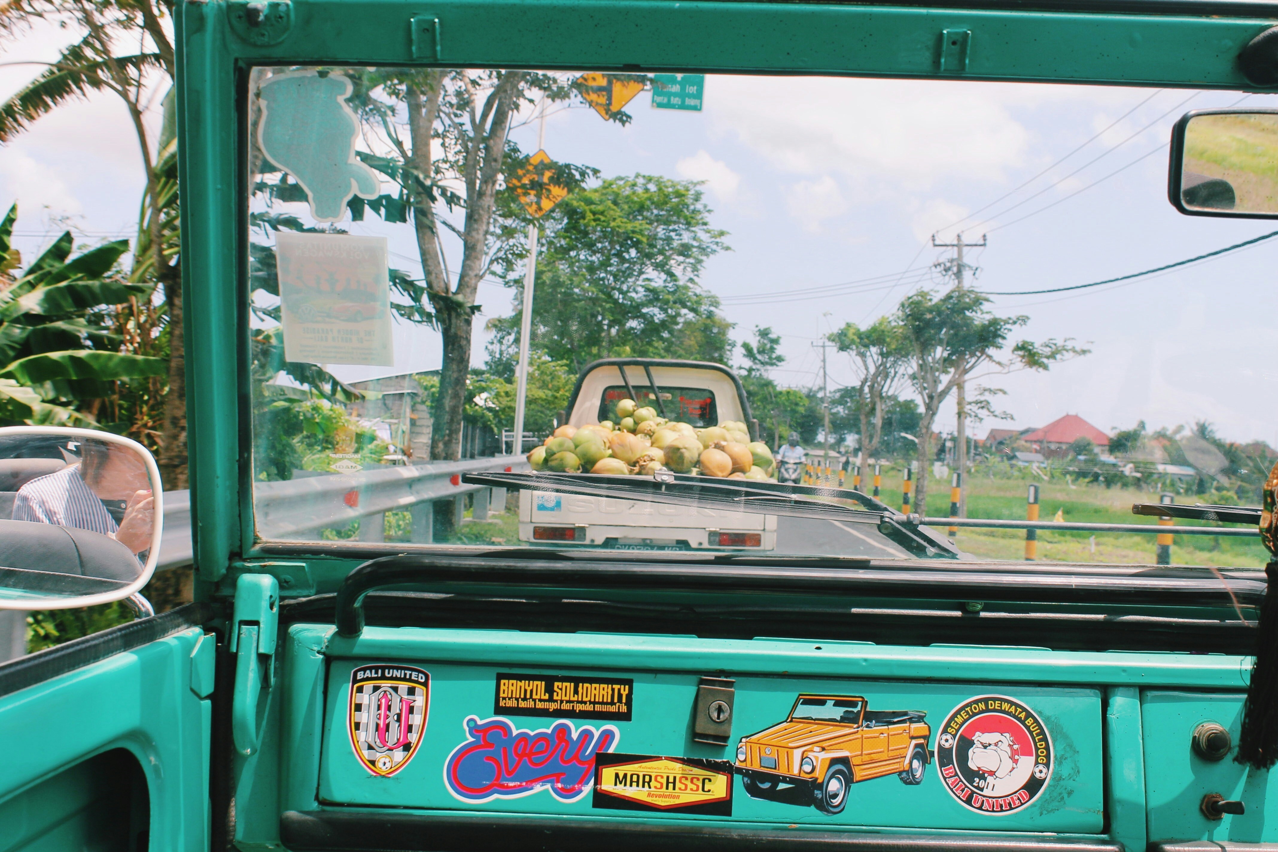 View from the driver's seat of a vintage vehicle, showcasing colorful stickers and a truck loaded with coconuts on a sunny day.
