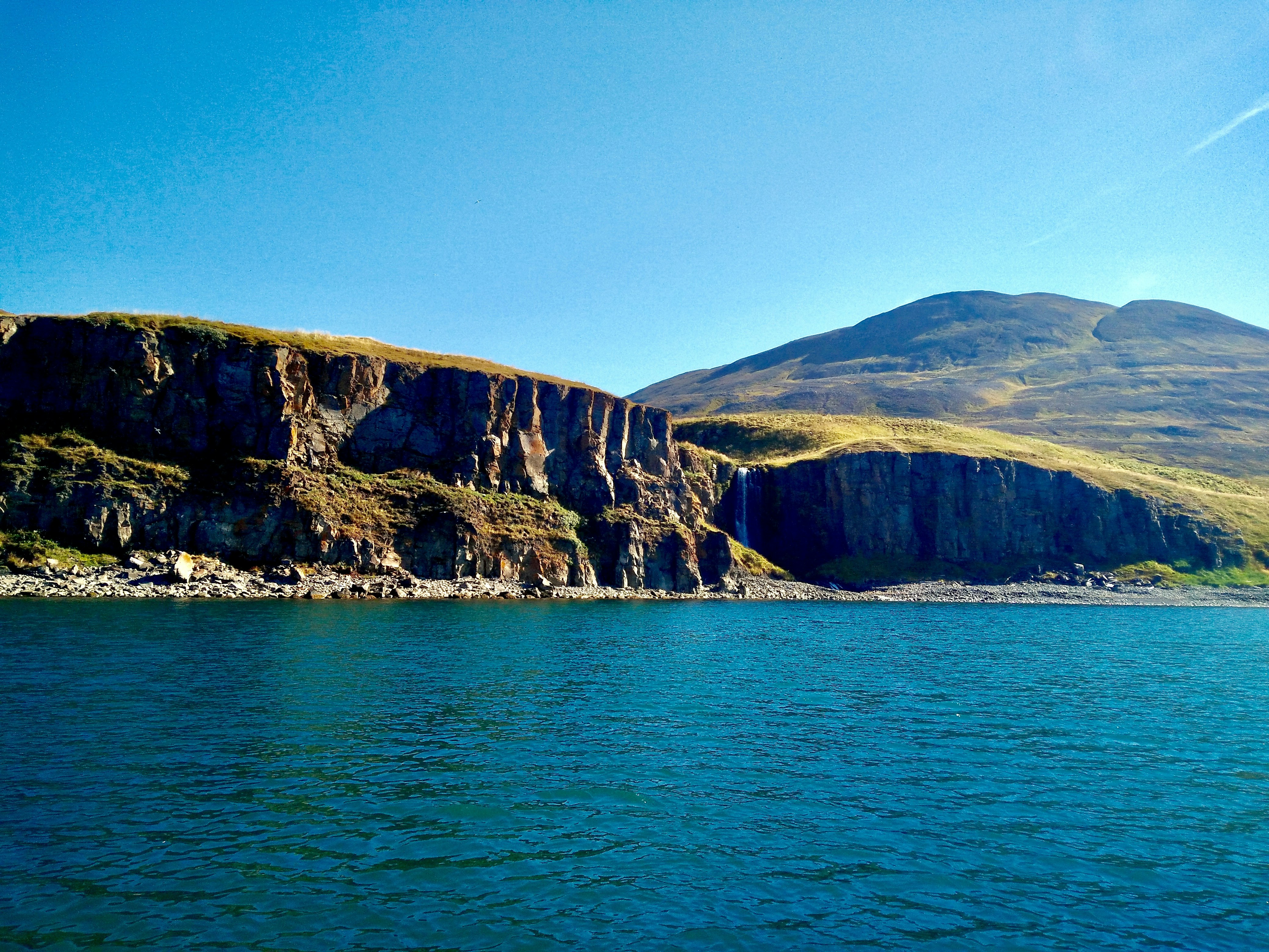 Lush cliffs rise dramatically above a serene blue lake, with a gentle waterfall cascading down the rocky face. The clear sky enhances the vibrant colors of the landscape.