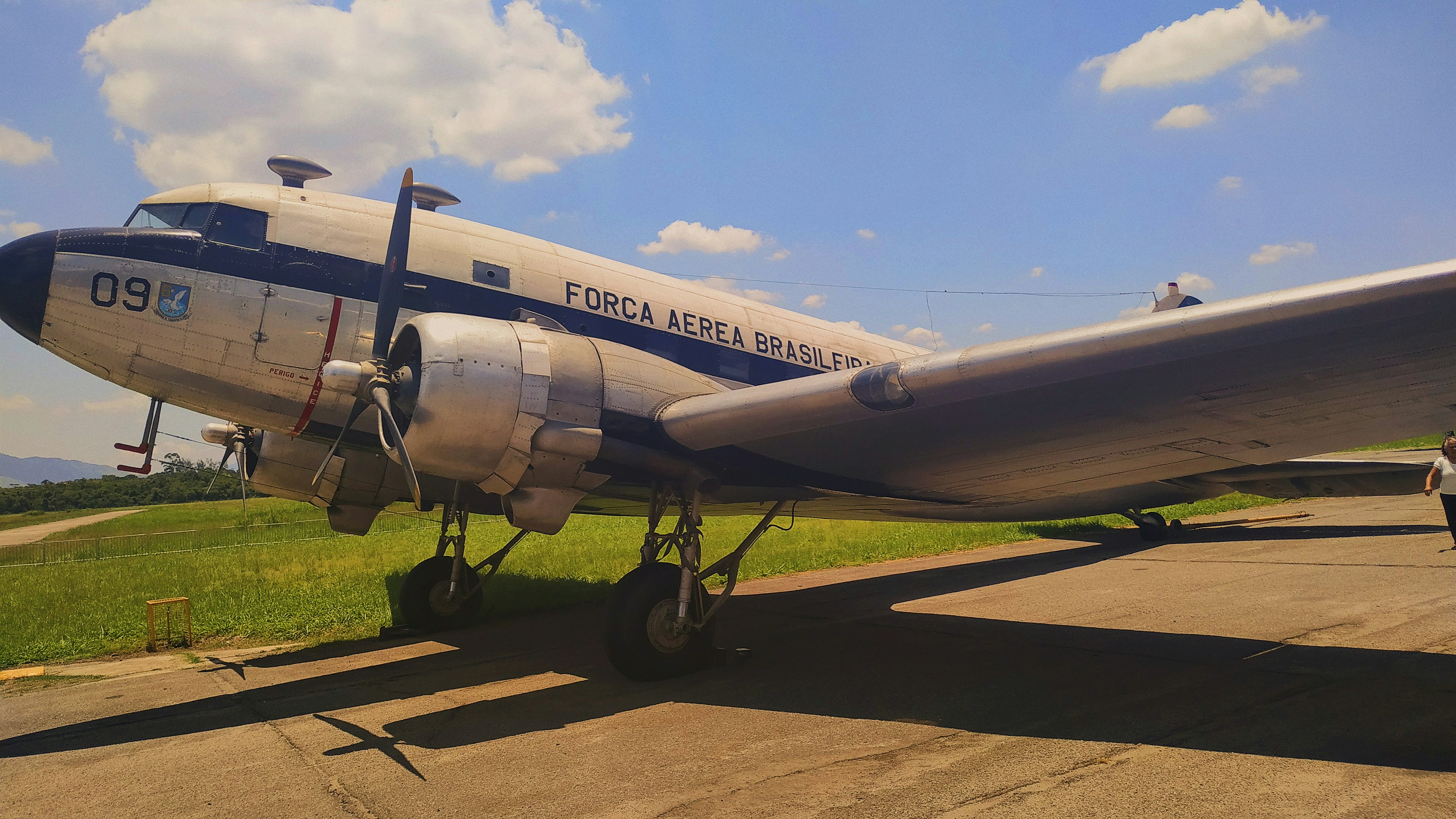 Historic propeller airplane resting on a sunlit tarmac with a backdrop of green grass and blue sky.