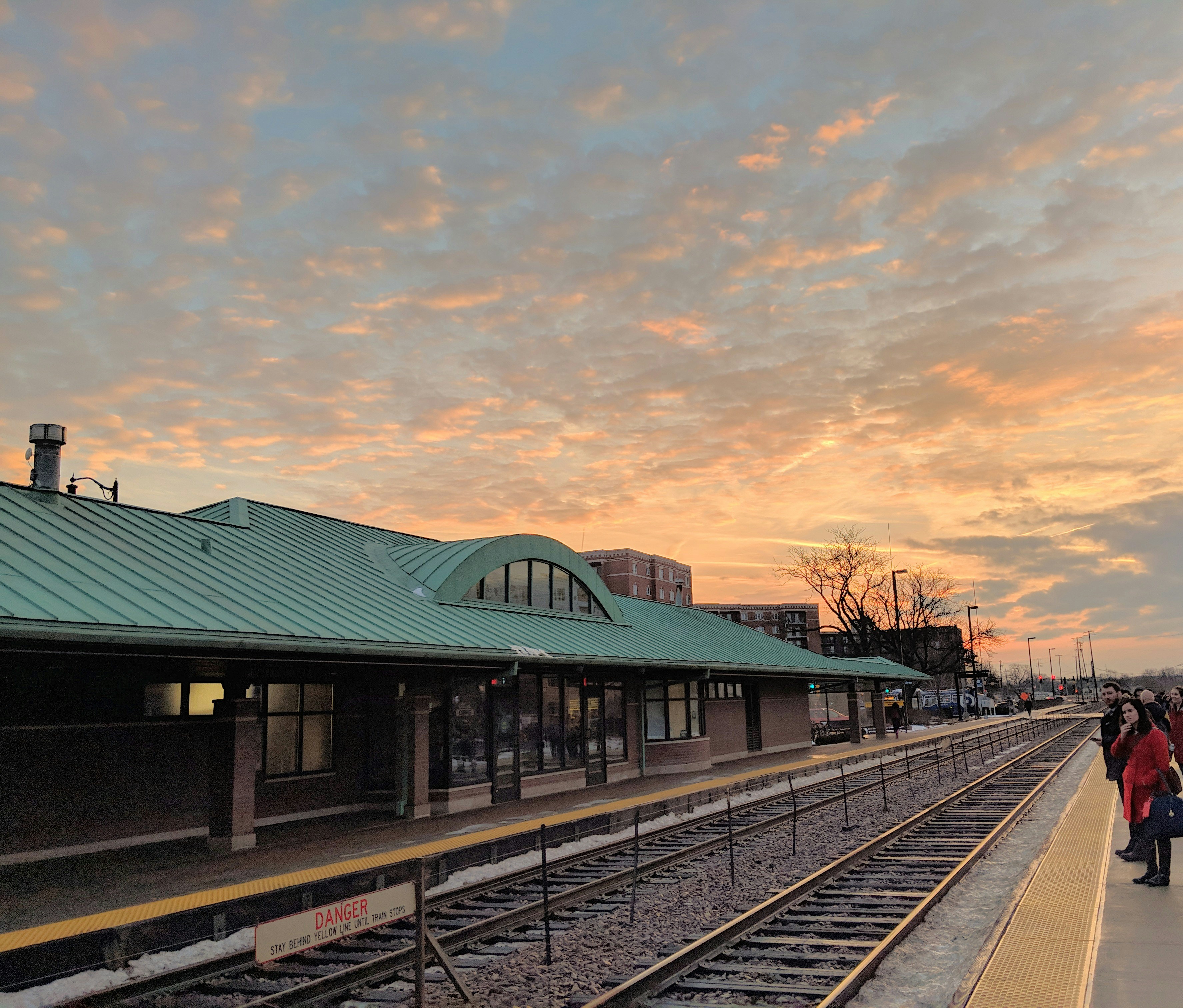 Train station under a colorful sunset, with silhouettes of waiting passengers along the platform. The scene captures the calm before the evening journey.