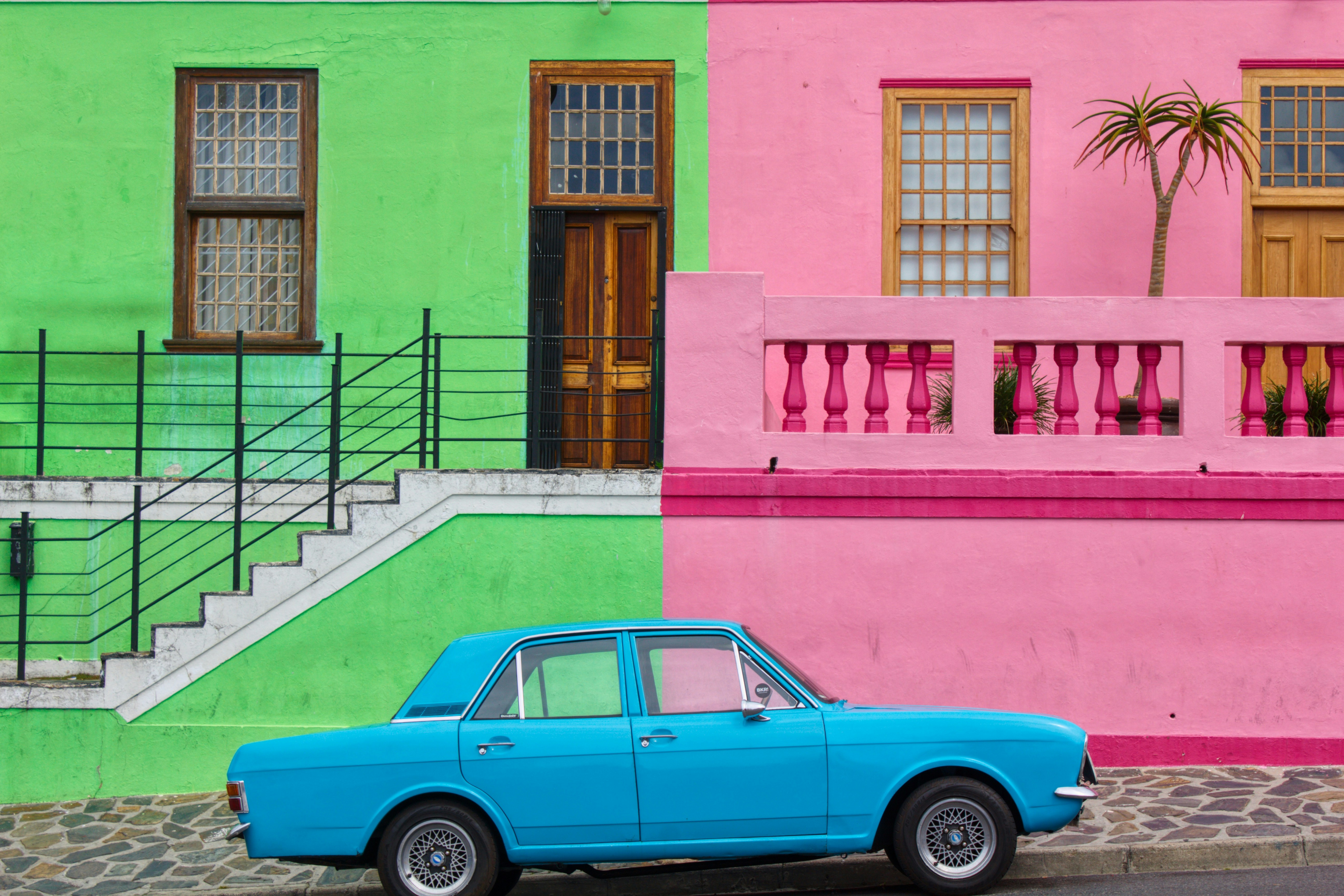 Blue vintage car parked against a vivid green and pink building with geometric lines.