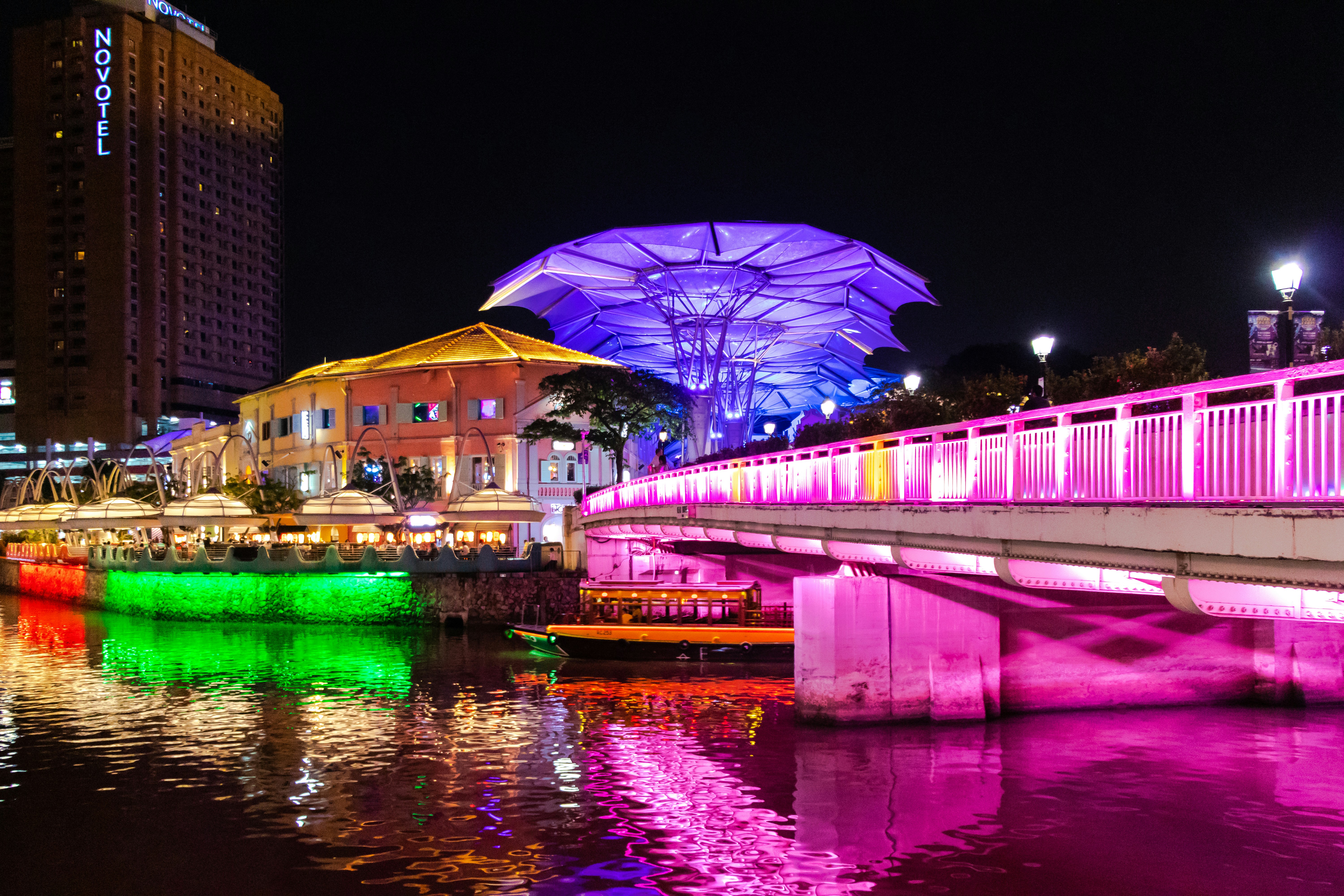 Vibrant lights illuminate the riverside, showcasing a colorful bridge and modern architecture under a night sky.