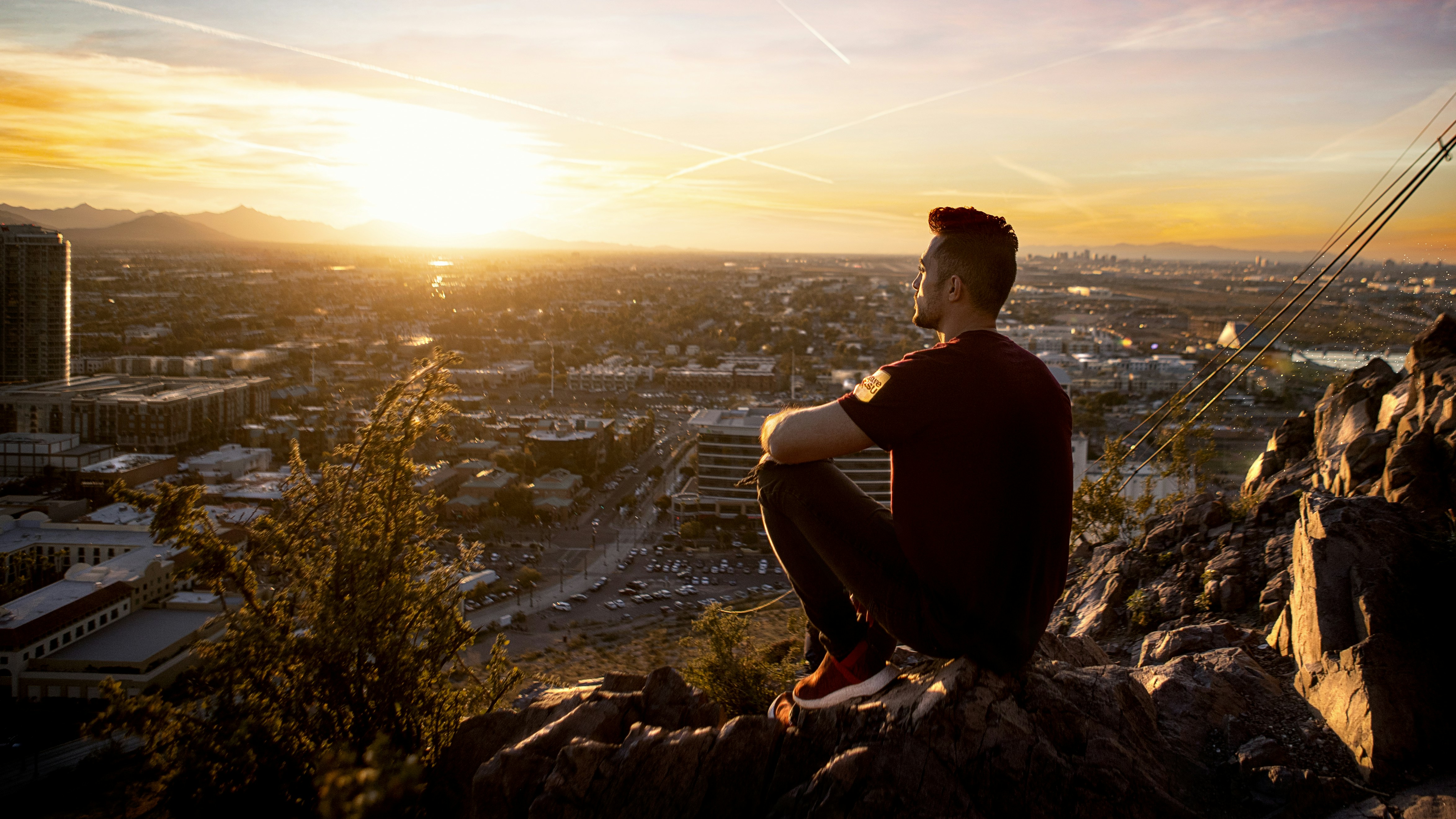 Silhouette of a man sitting on a rock overlooking a sprawling cityscape at sunset.