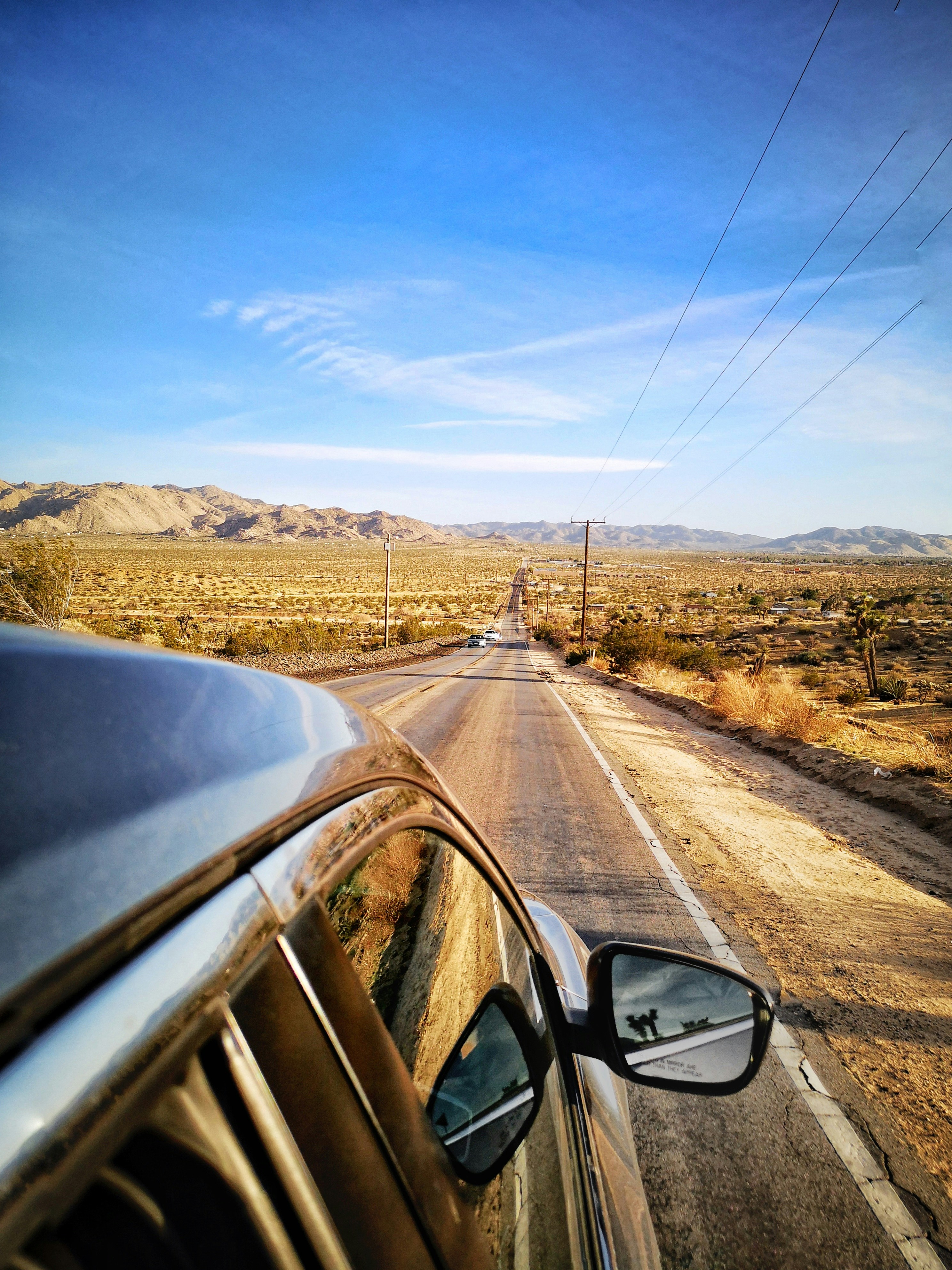 Desert highway stretching into the horizon, framed by rugged mountains and sparse vegetation, with a vehicle's side mirror reflecting the vast landscape.