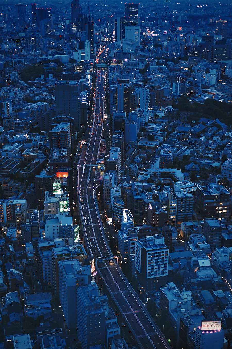 Aerial view of a bustling cityscape at night, showcasing illuminated streets weaving through dense buildings. The vibrant lights contrast against the deep blue night sky.