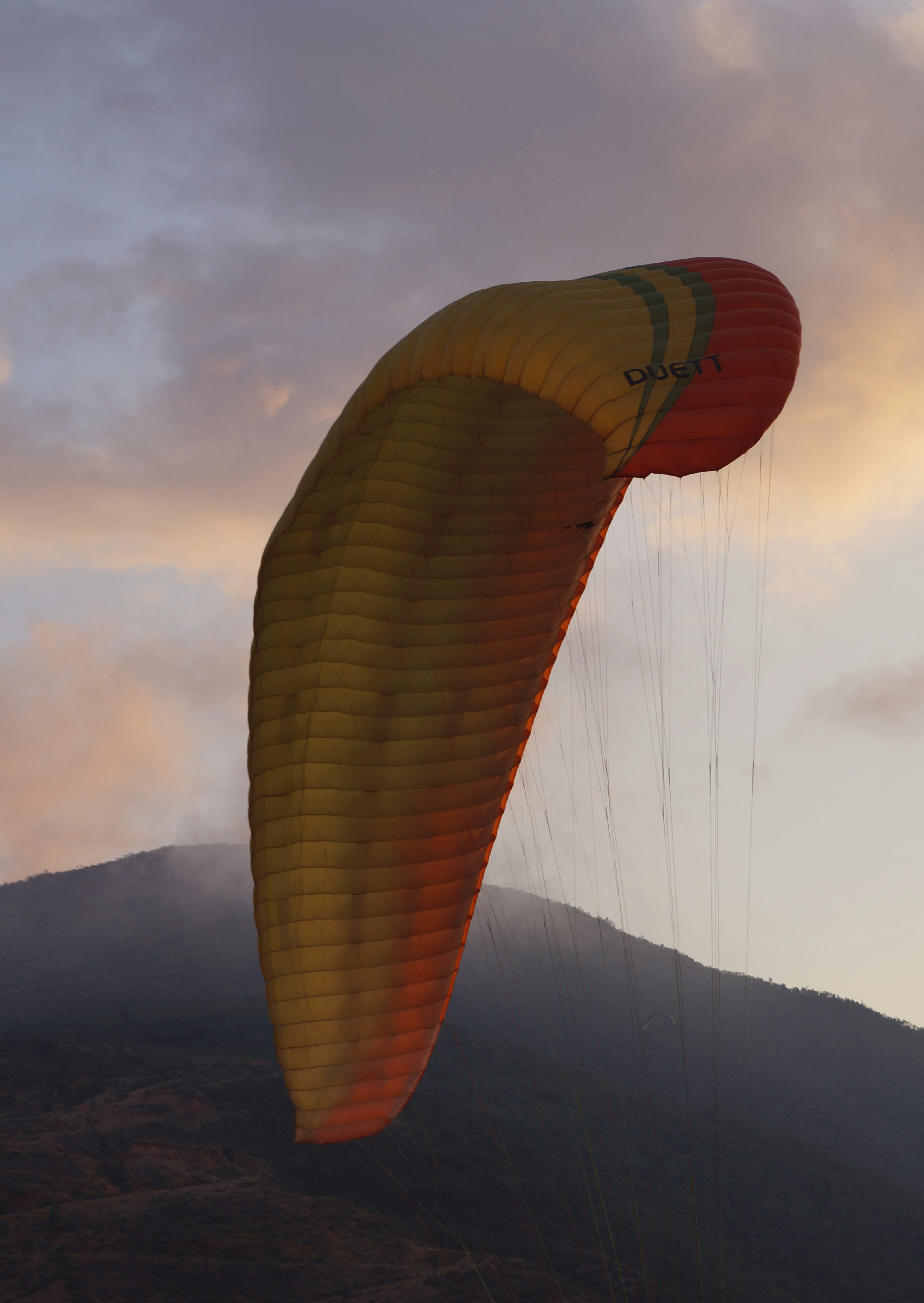 A vibrant yellow and orange paraglider suspended against a backdrop of soft clouds and a distant mountain range during sunset.
