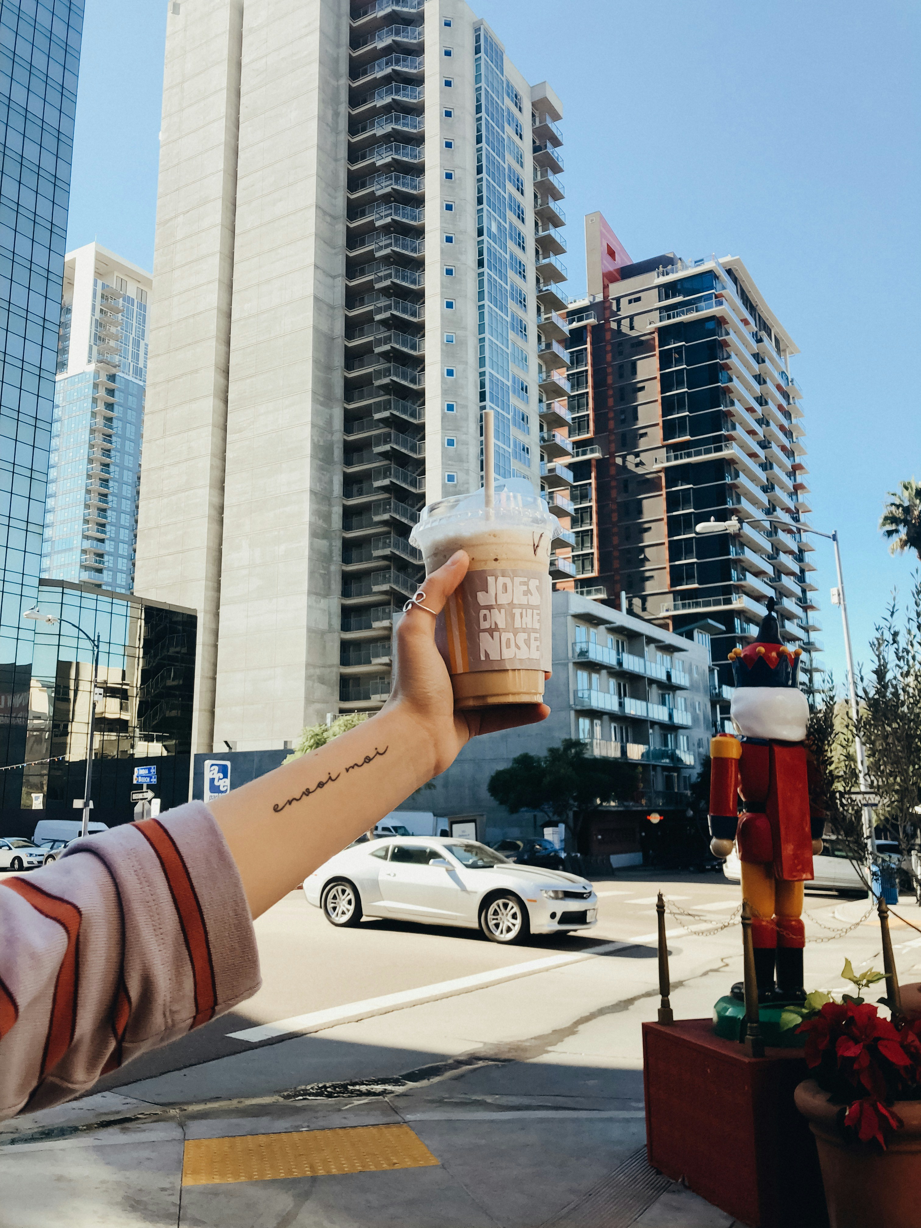 A hand holds a coffee cup with the label 'JOES ON THE NOSE' against a backdrop of modern skyscrapers and urban architecture. A nutcracker figure adds a festive touch to the scene.