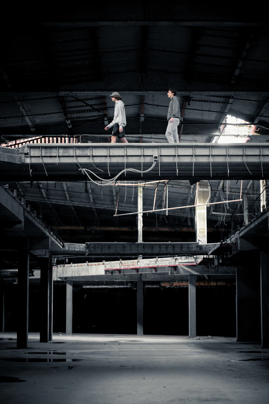 Two figures walk a suspended catwalk inside a dim, abandoned factory—an architectural photograph capturing isolation and decay.