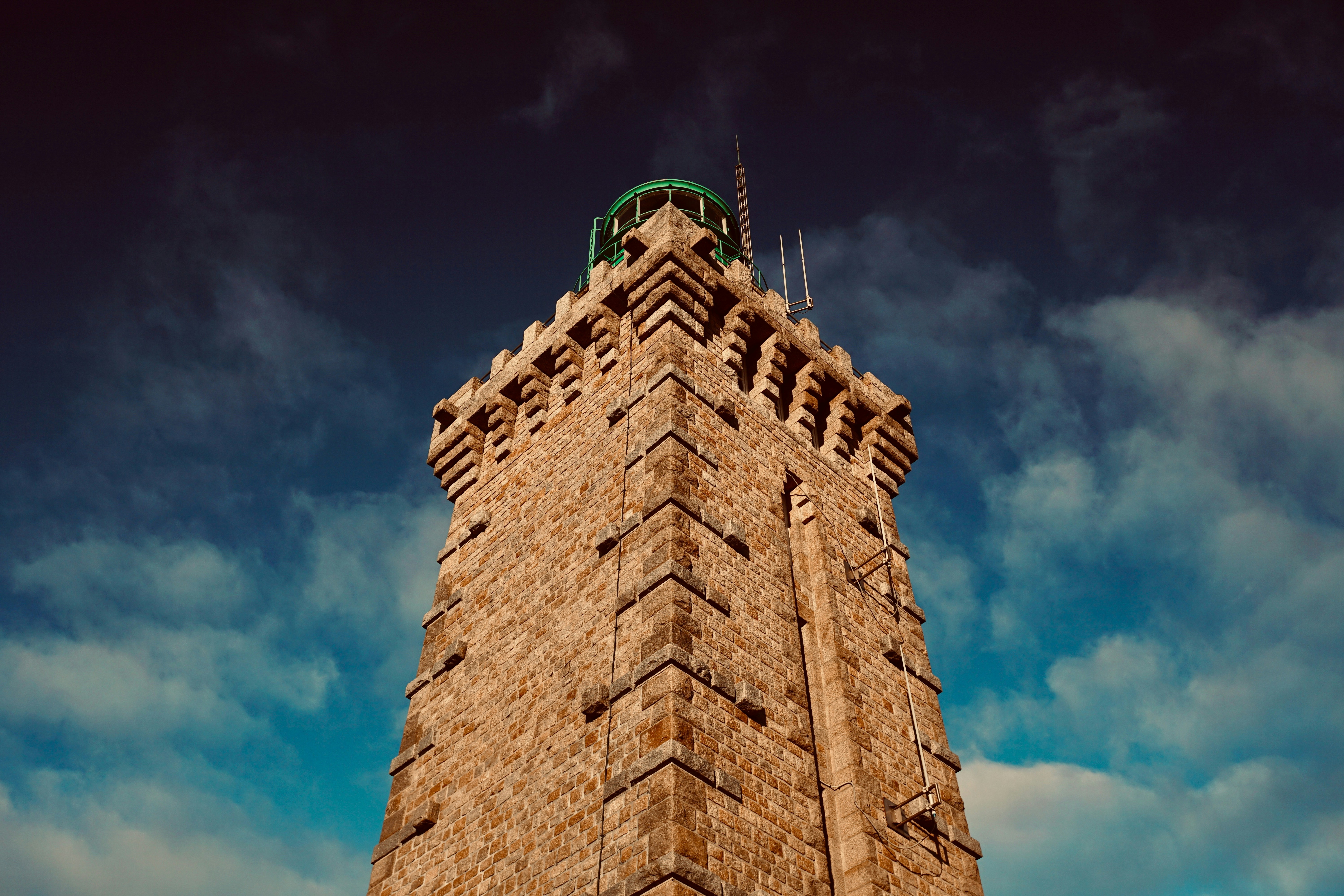 Historic stone lighthouse standing tall against a backdrop of dramatic clouds. The structure features a green lantern atop its tower.