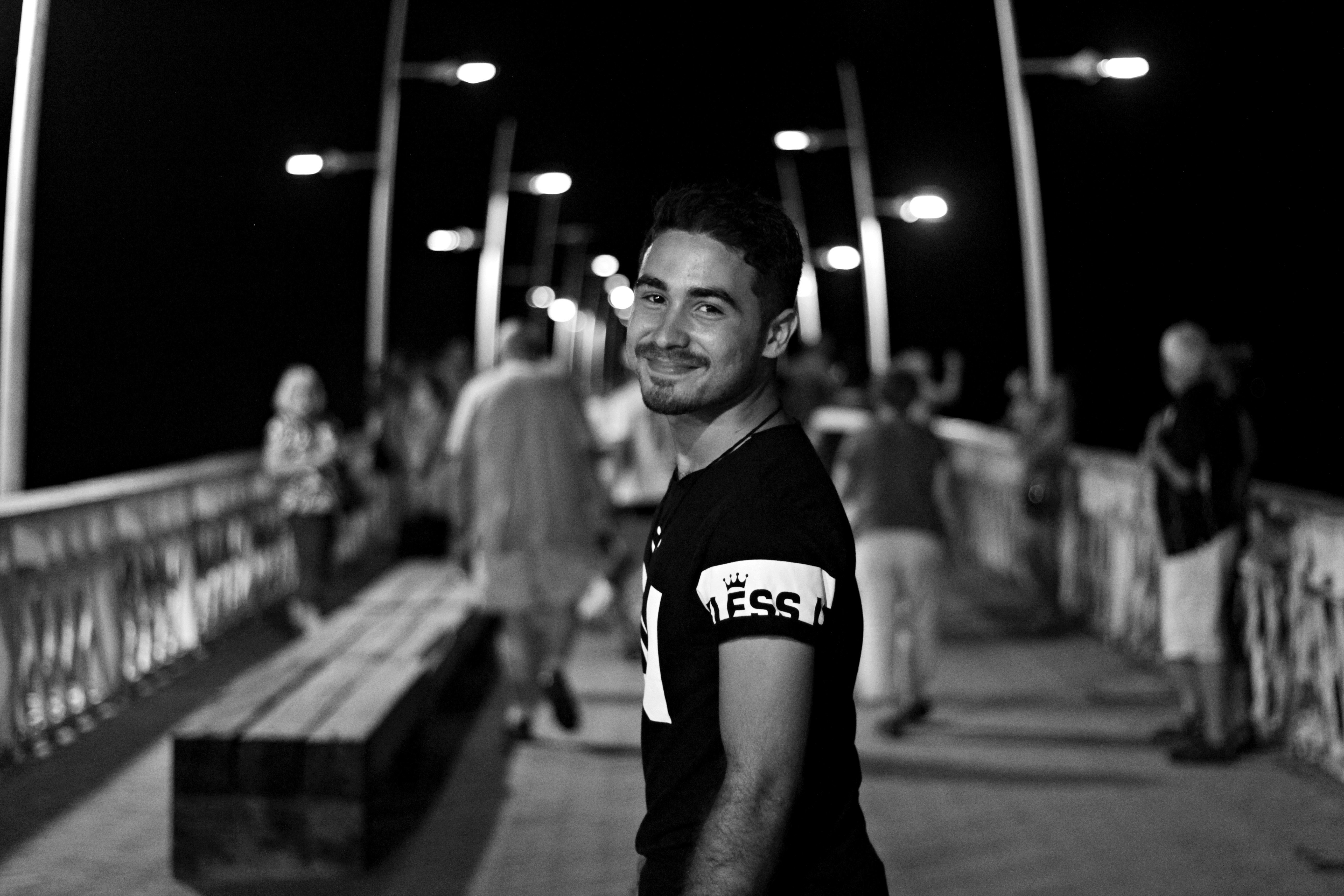 Young man smiling while walking on a busy boardwalk at night, with blurred figures in the background creating a sense of movement.